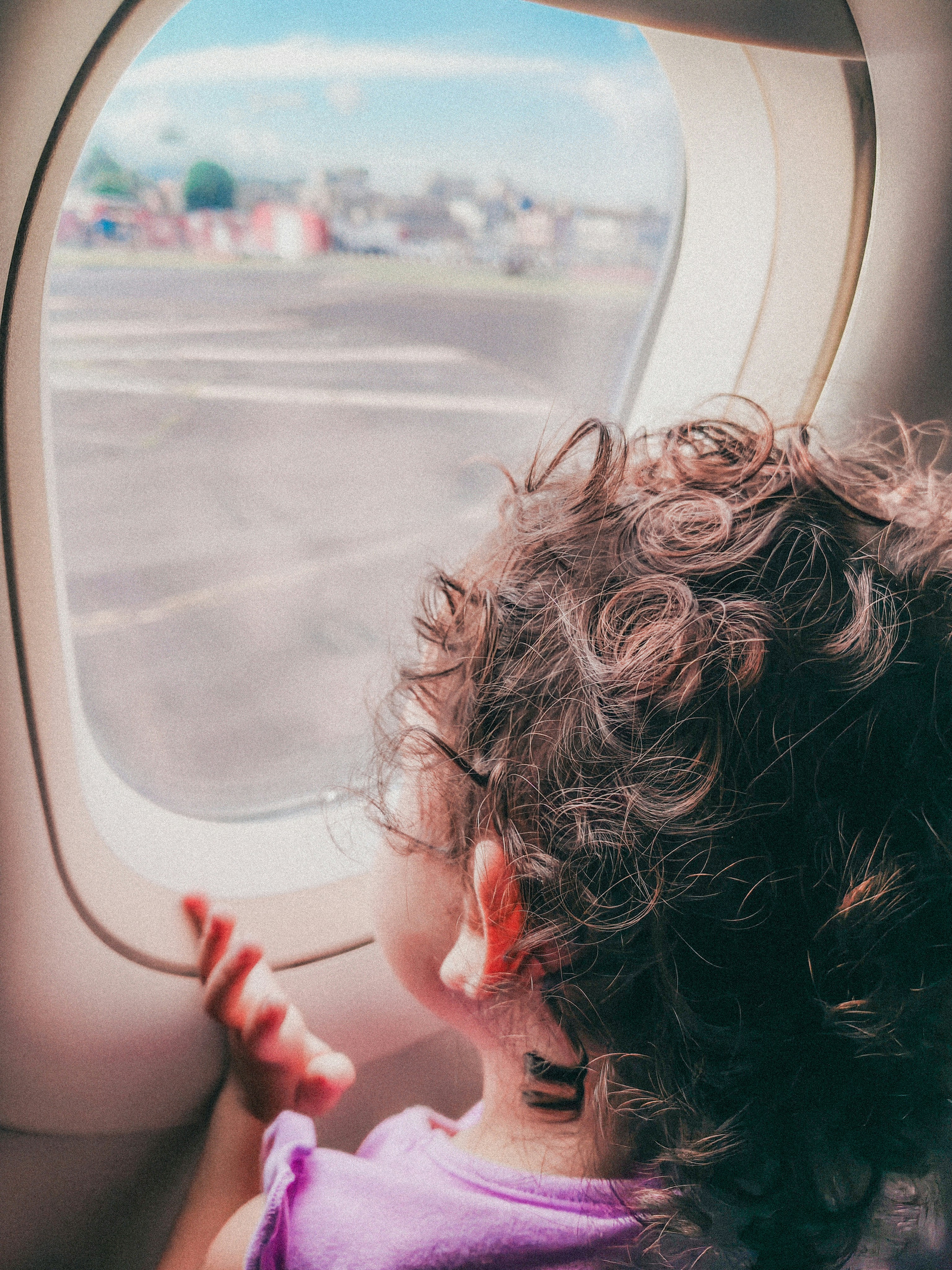 A child with curly hair looking out an airplane window at an airport tarmac