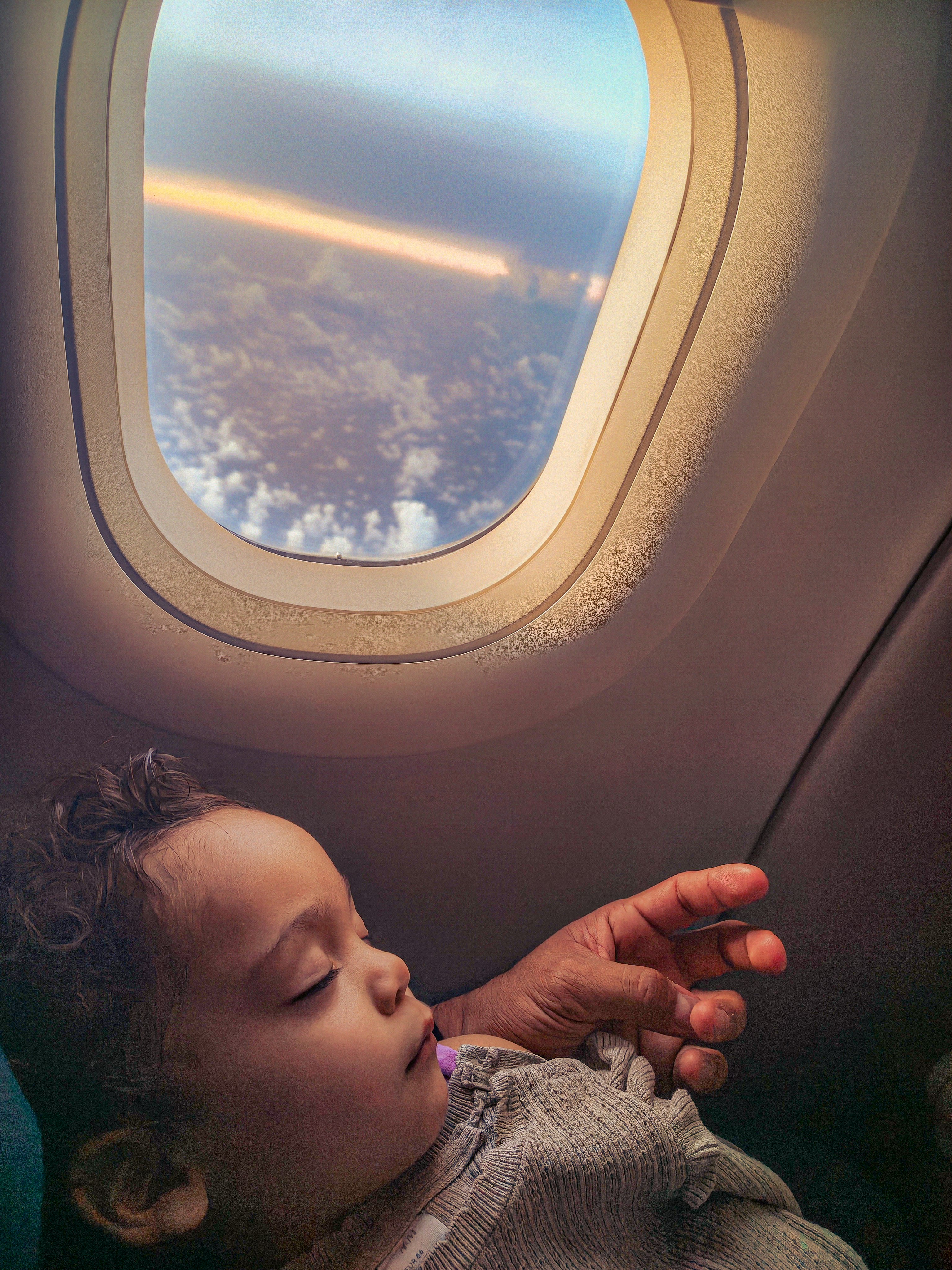 A child sleeping against an adult hand by an airplane window with clouds below