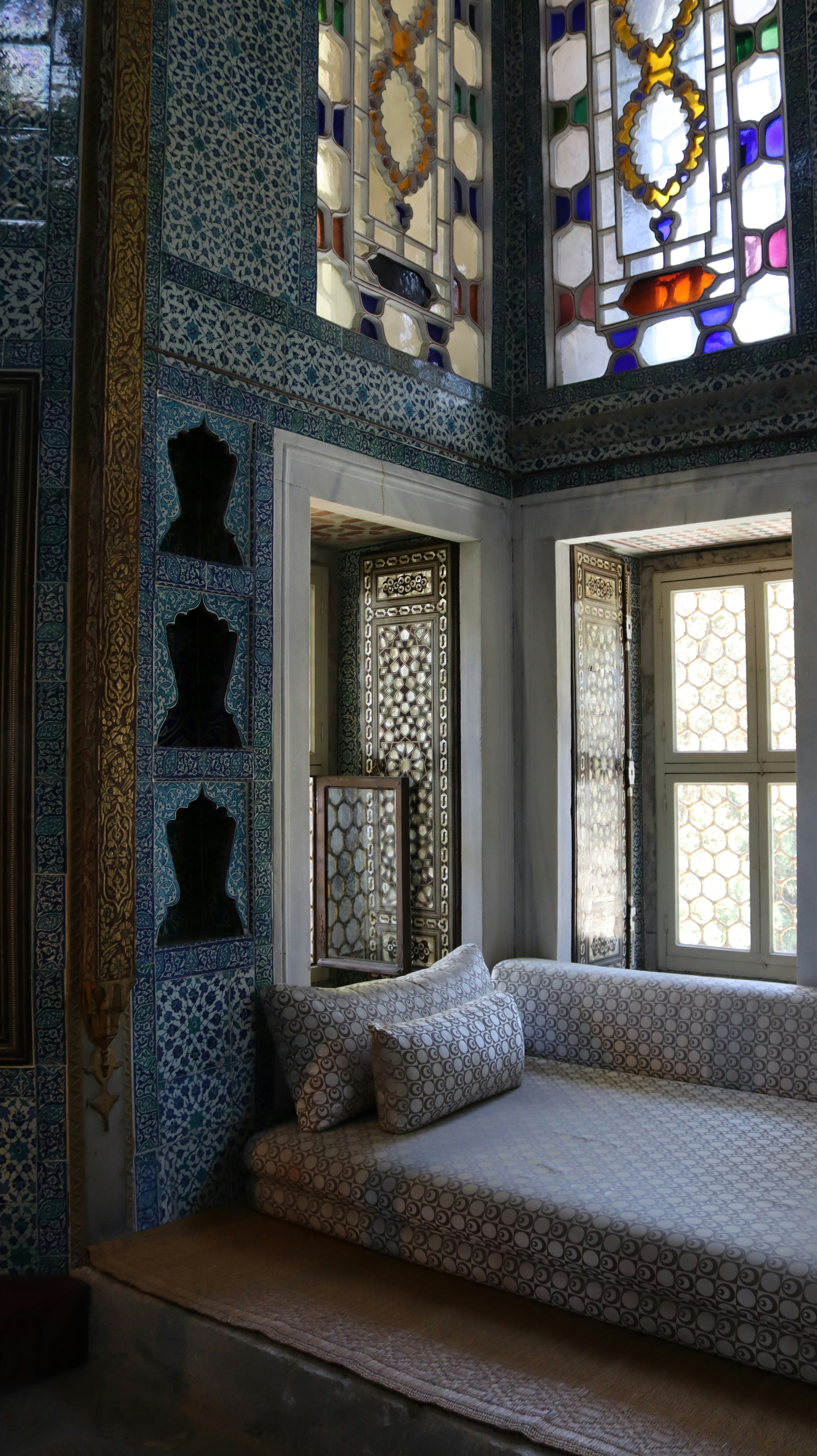 Intricate blue-and-white tiled walls frame a sunlit window seating nook with patterned cushions and ornate stained glass overhead. A tranquil, domestic space showcasing craft details and soft natural light.