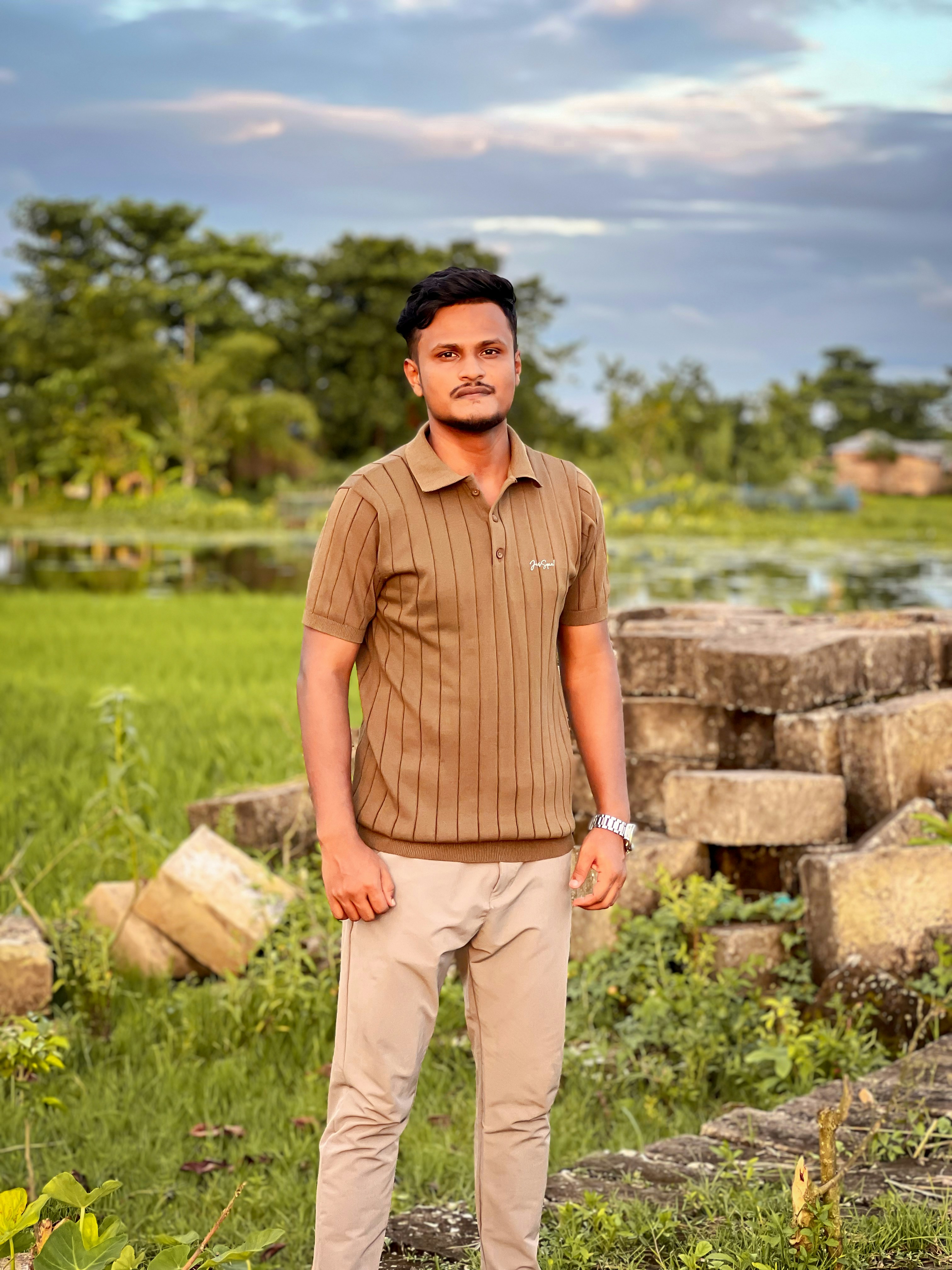 Man in brown polo and light pants stands near stone blocks by a water body with greenery and a soft sky behind.