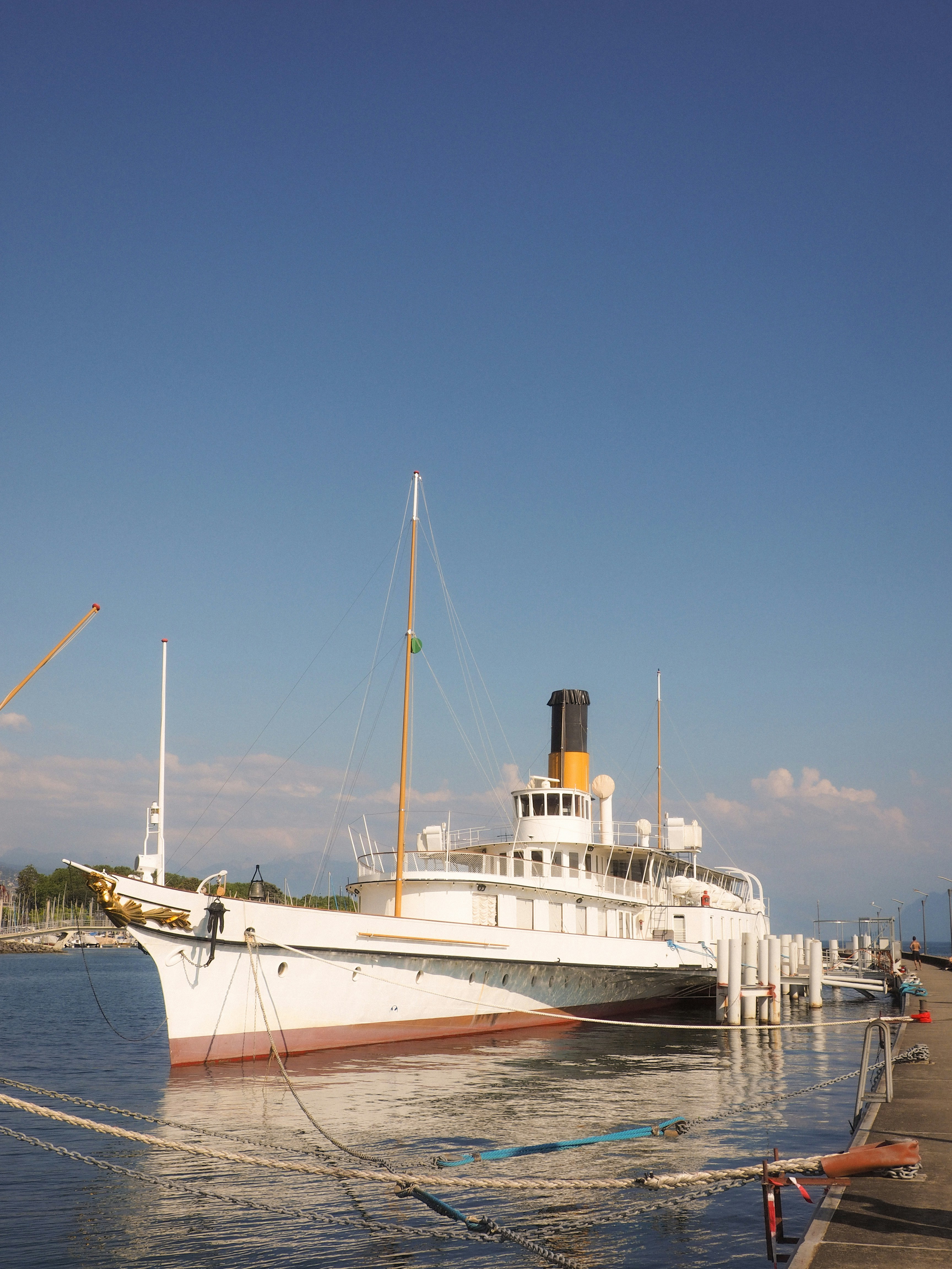 Classic steamship moored at a calm harbor with a pier and mooring ropes; a tranquil maritime scene emphasizing the vessel's white hull and tall smokestack.