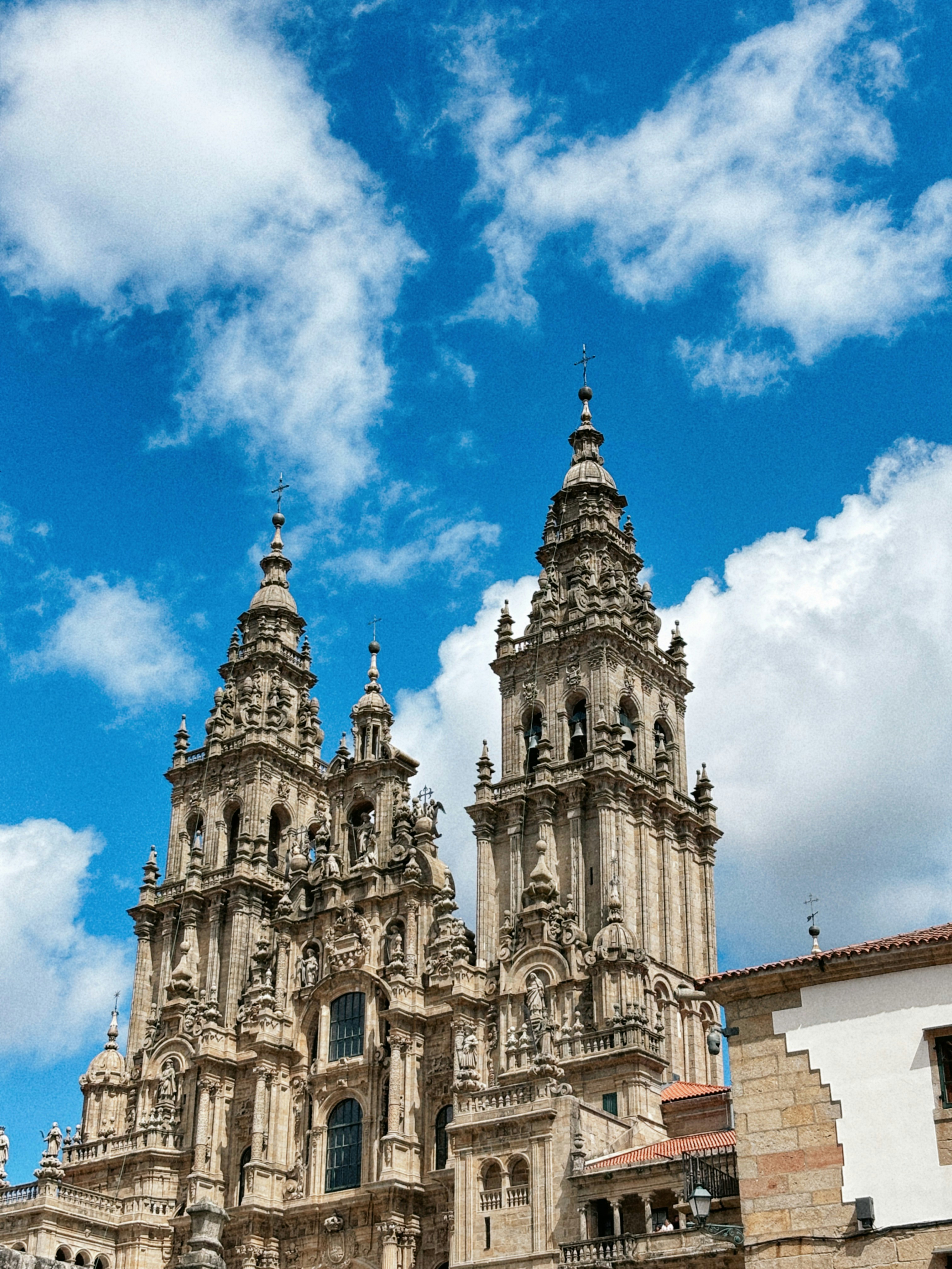 Gothic-style cathedral towers rise against a bright blue sky with scattered clouds, showcasing intricate stonework and architectural detail.