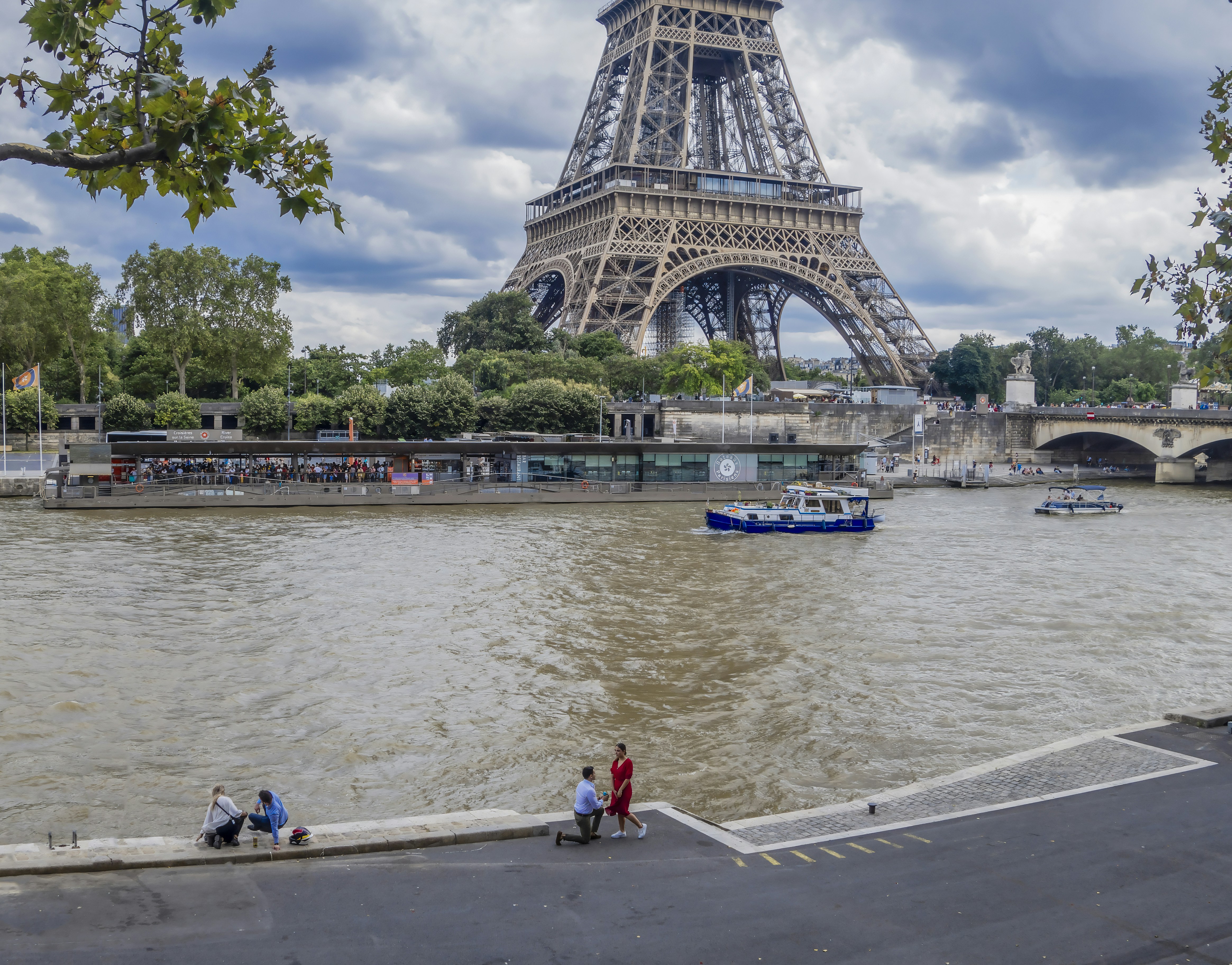 City riverbank scene with the Eiffel Tower in view and people by the water, capturing a leisurely moment along the Seine.