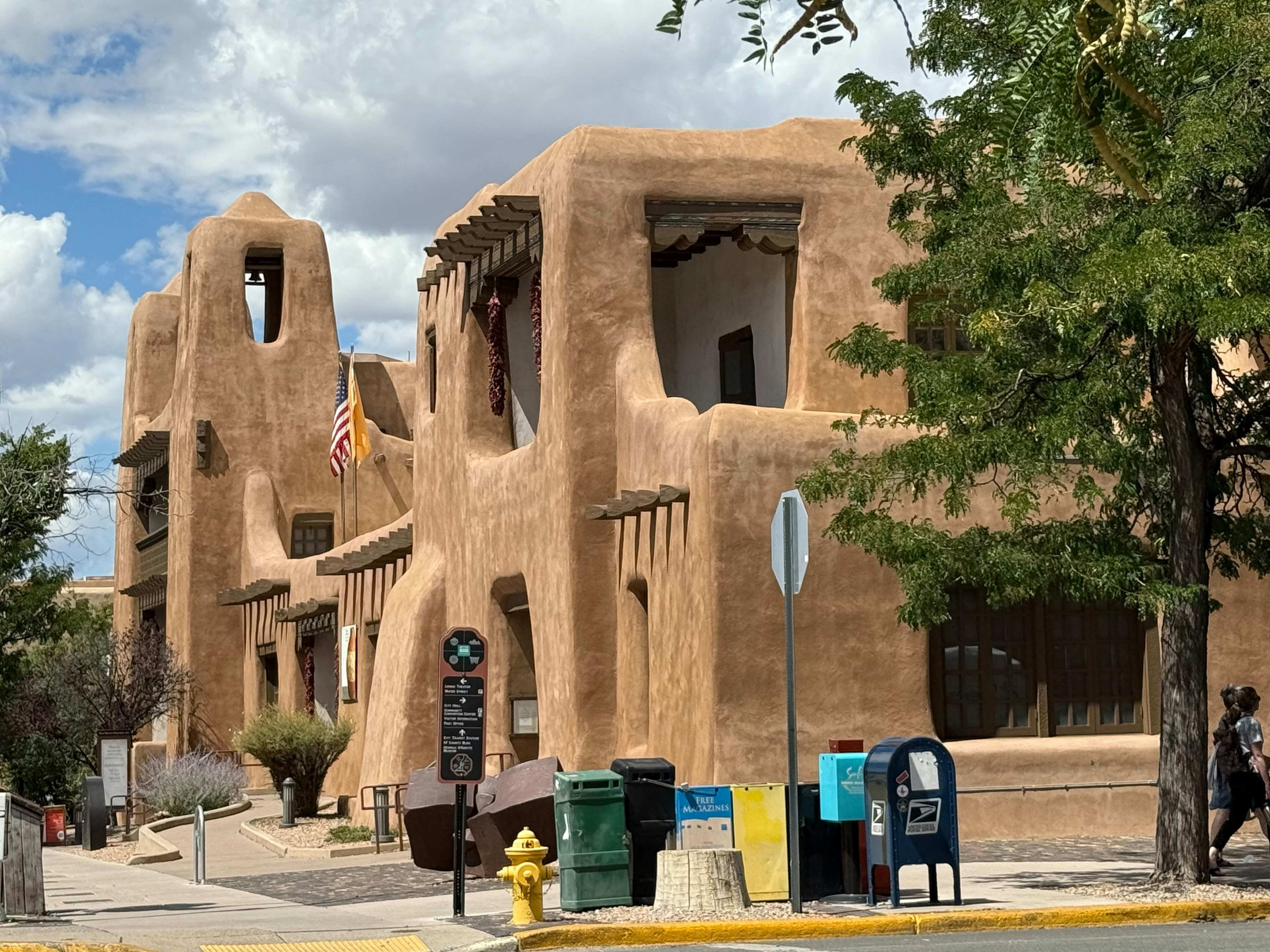 Adobe-style building with rounded towers and a street scene featuring mailboxes and a parked car, conveying a small-town southwestern atmosphere. Unknown logo signage visible near the facade.