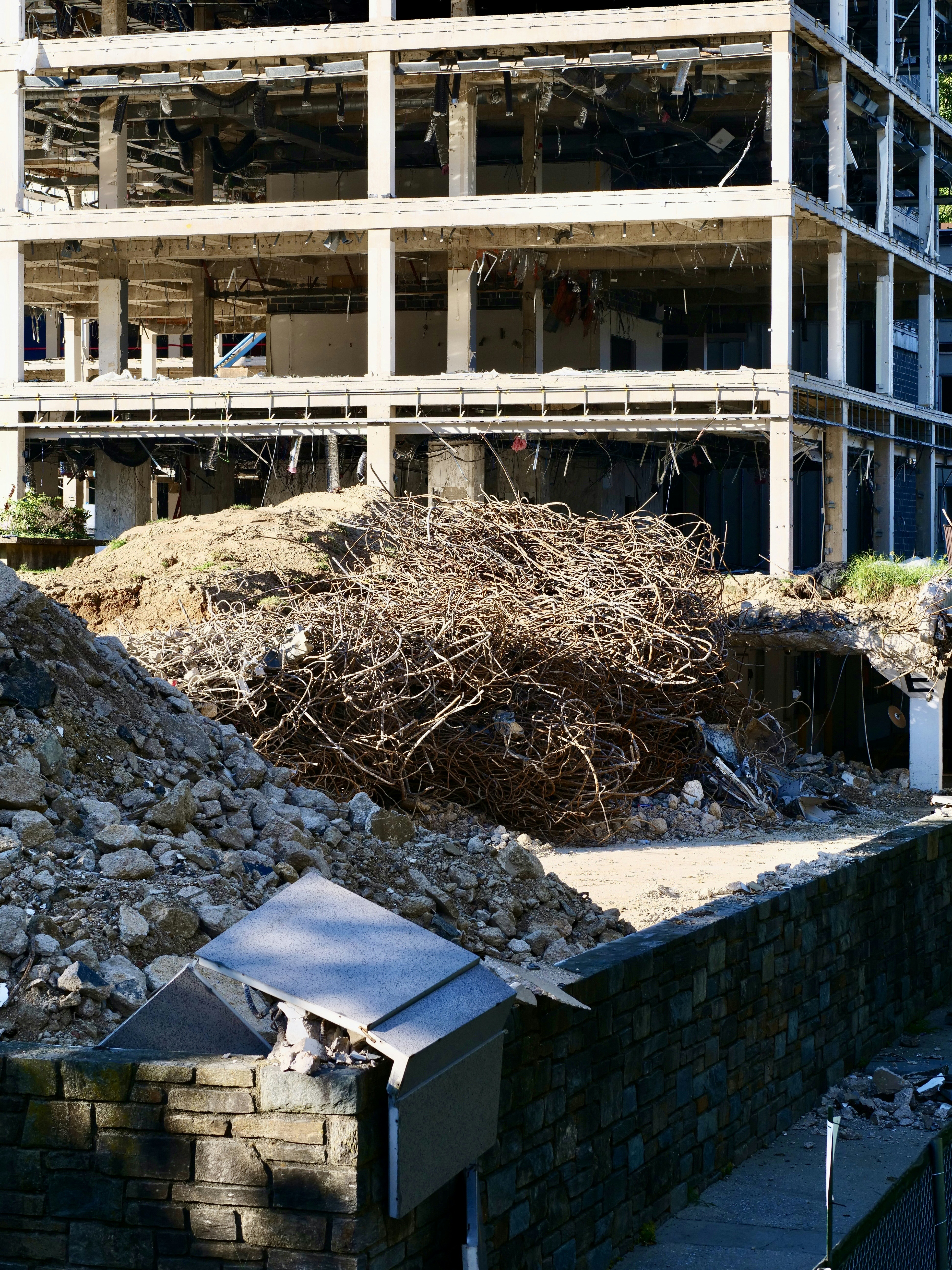 Under-construction building with exposed concrete floors and a tangle of rusted rebar in the foreground; a pile of dirt and debris sits along a brick retaining wall, with a dark, partially lit interior visible through the skeletal structure.