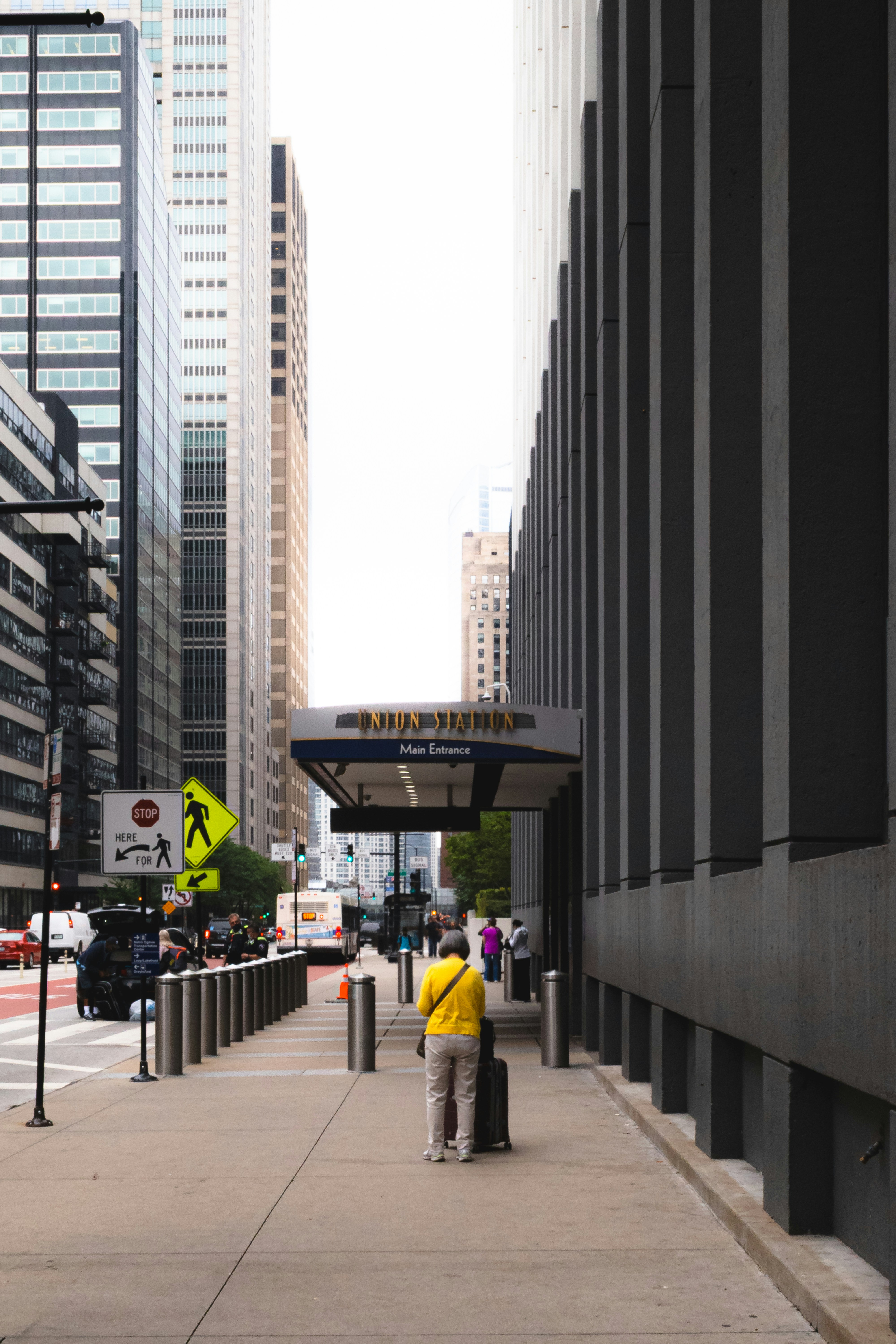 Traveler in a yellow raincoat with rolling suitcase stands under a city canopy between tall buildings, guiding the viewer toward the urban entrance. City street scene with pedestrians and vehicles.