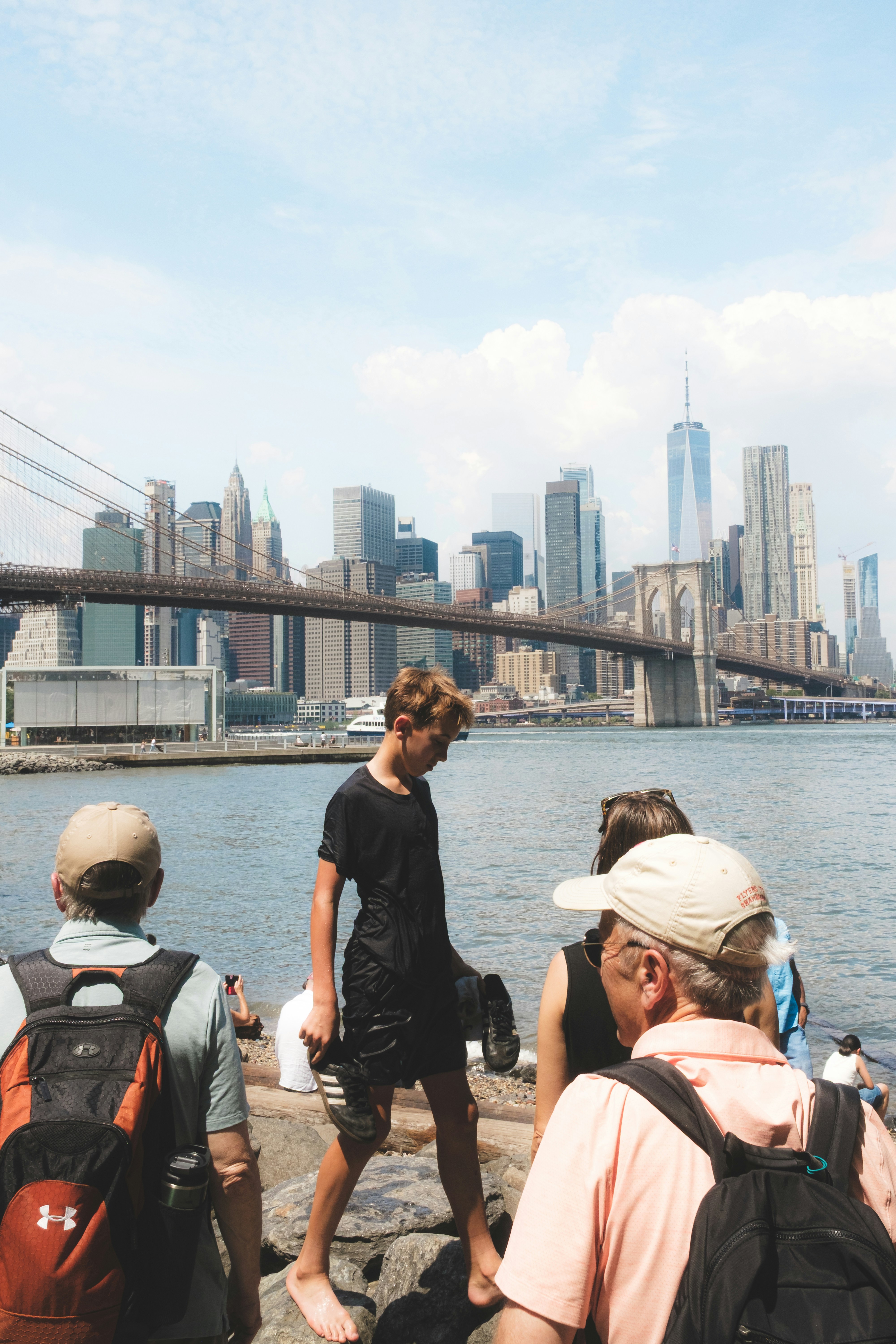 A group of people stands near the riverfront with a young teen stepping onto rocks as Brooklyn Bridge and skyline loom in the background. A travel scene capturing interaction and urban backdrop.