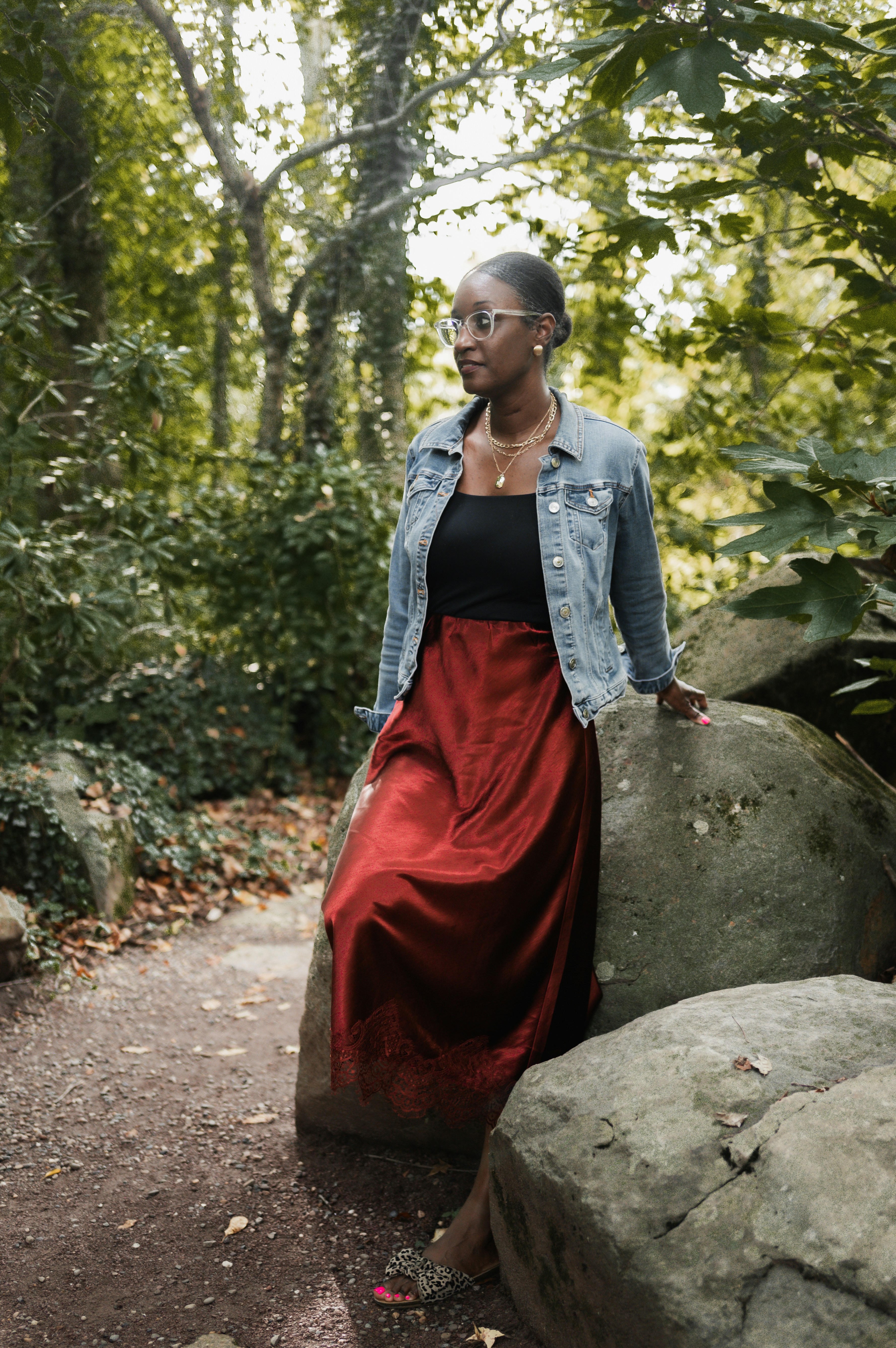 Woman stands among large rocks in a sun-dappled garden, wearing a denim jacket, black top, and red skirt; natural surroundings form a calm, portrait-oriented scene.