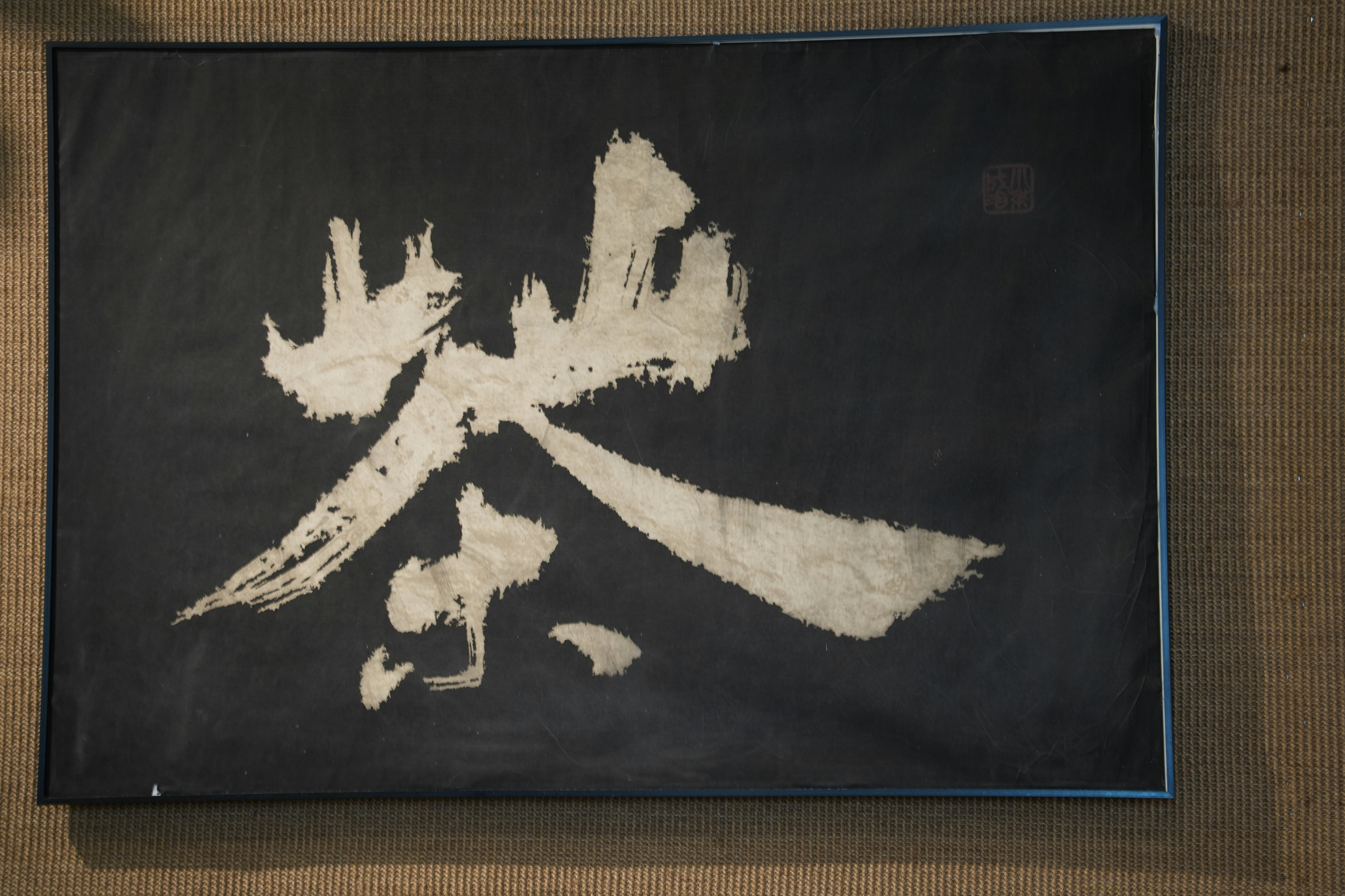 Bold white brushstroke characters on a dark canvas, framed and mounted on a textured wall. A logo-like piece emphasizing traditional calligraphy style.