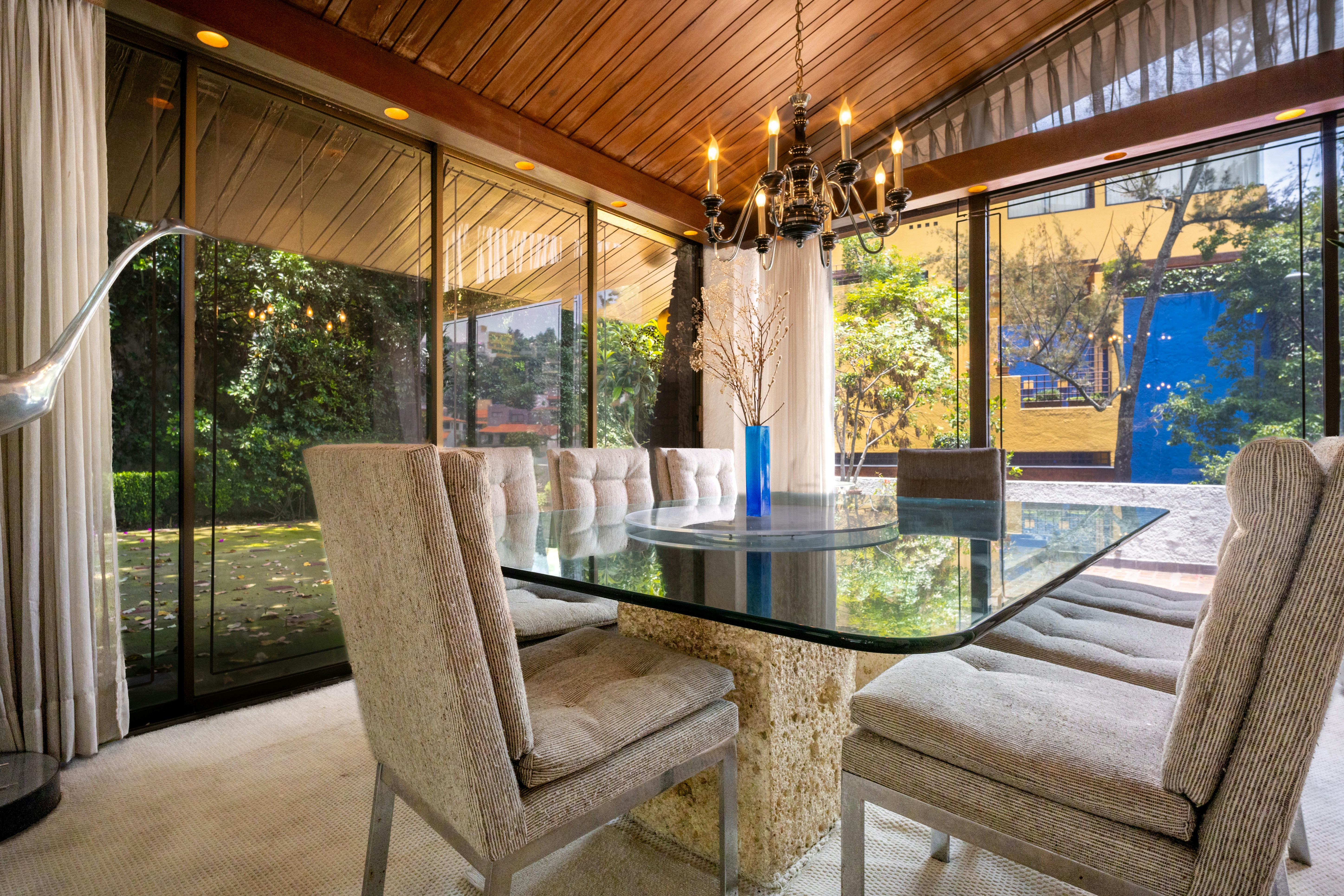 A modern glass-top dining table in a sunlit conservatory, surrounded by cushioned chairs and a chandelier, with floor-to-ceiling windows revealing an outdoor garden.