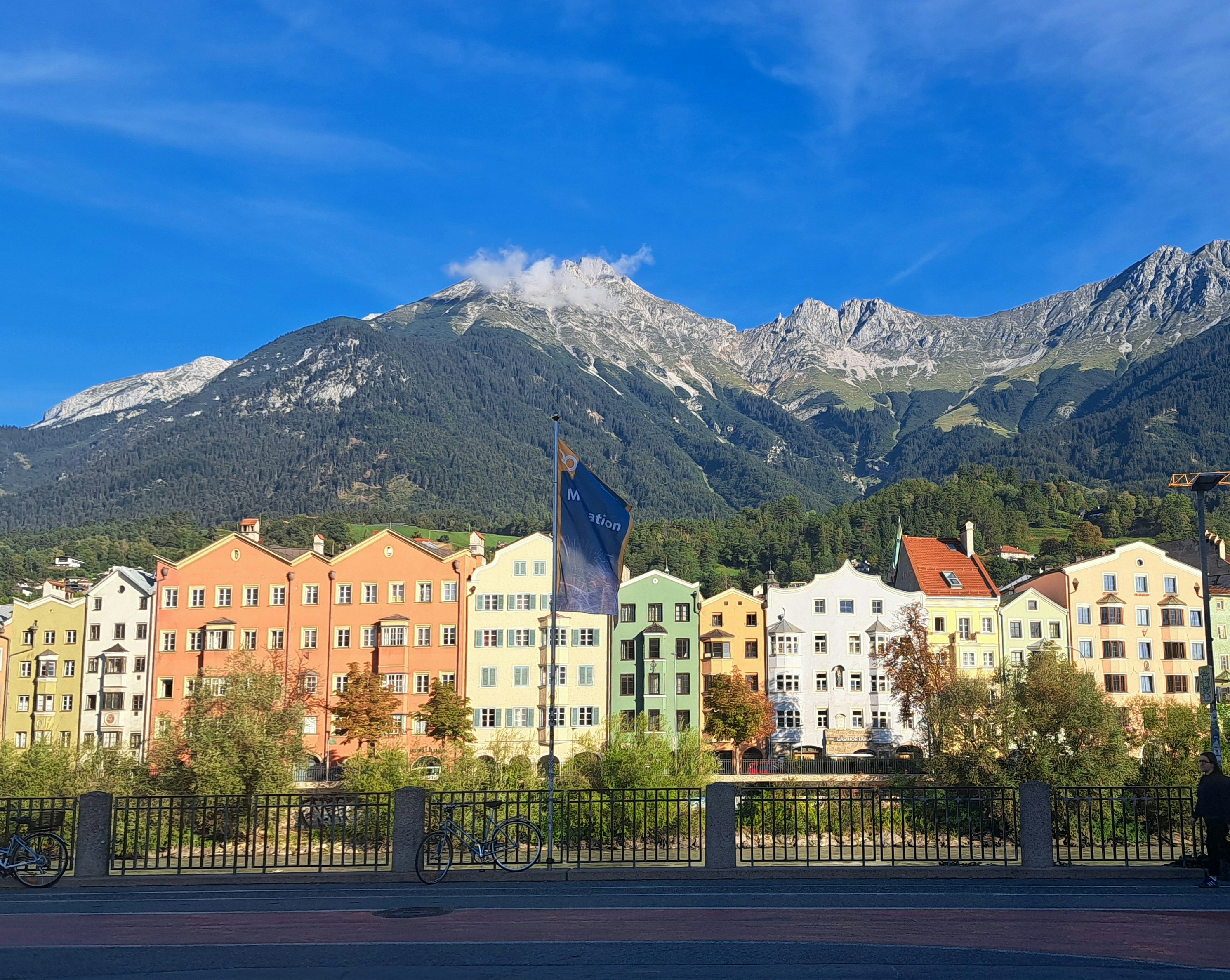 Colorful cottages line a riverfront with a railing and trees; alpine peaks loom in the background, under a clear blue sky.