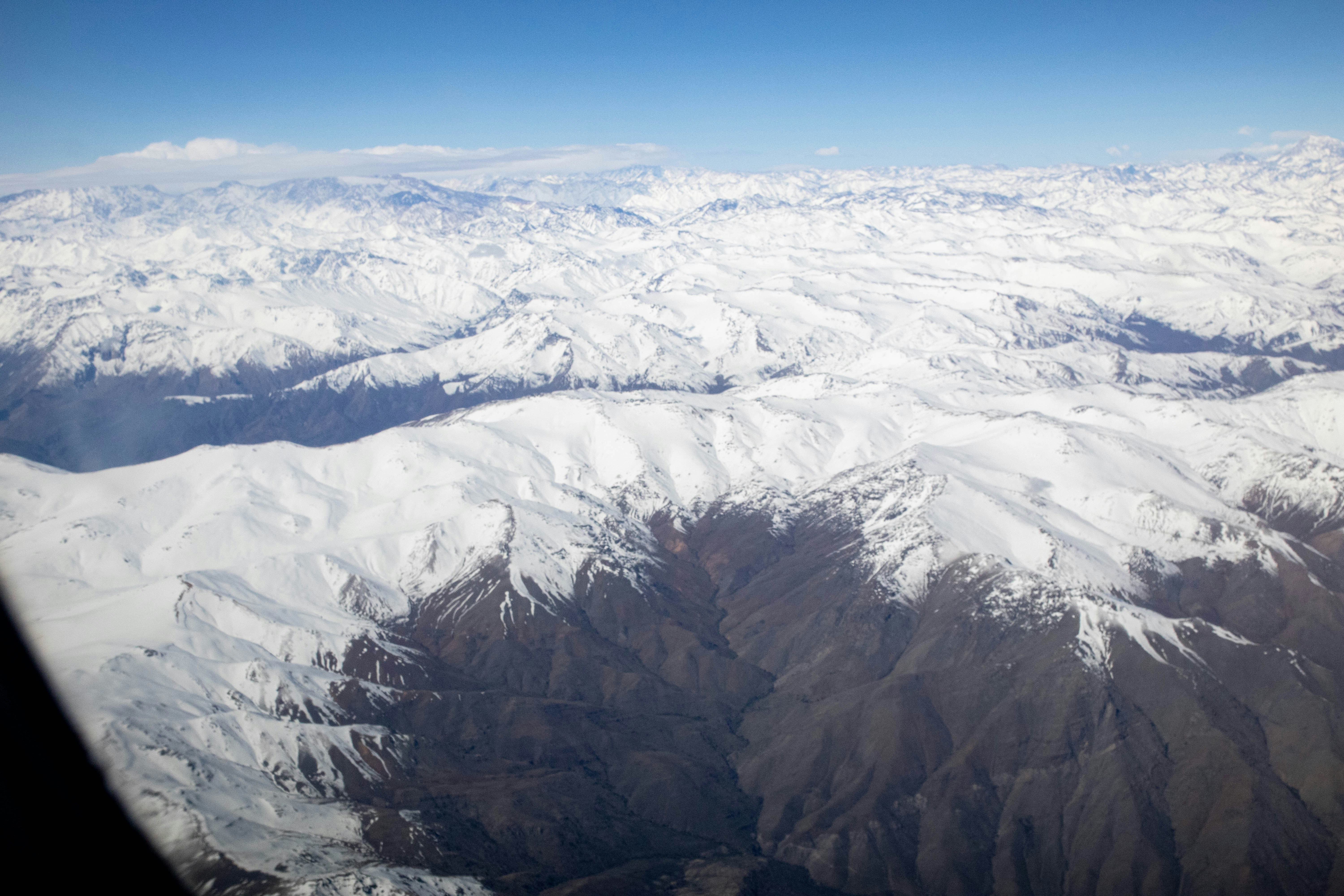 Snow-covered mountain range seen from an airplane window, with dark valleys below and a clear blue sky ahead.
