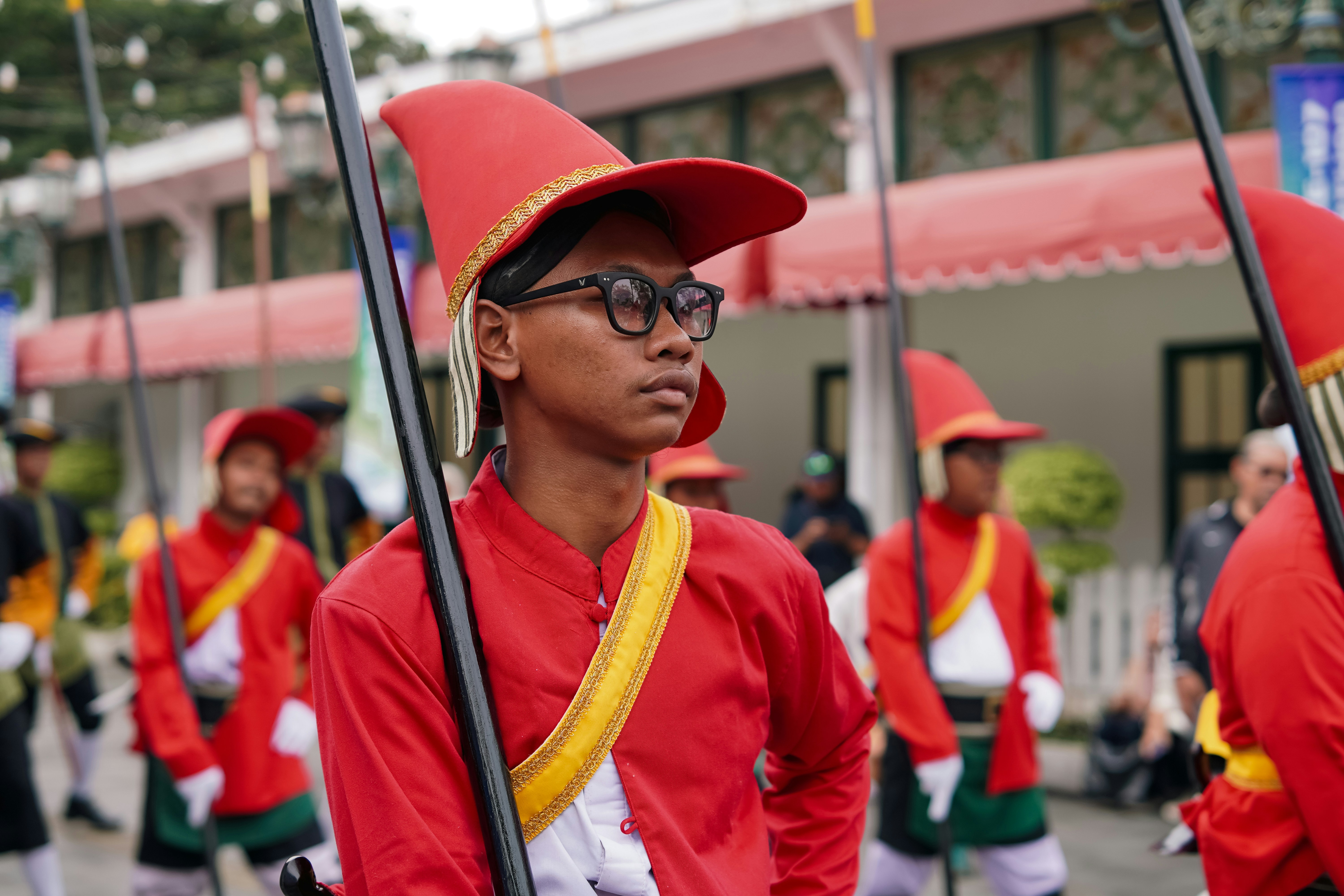 Cropped portrait of a young performer in a red uniform and hat, holding a baton during a parade with others in the background.