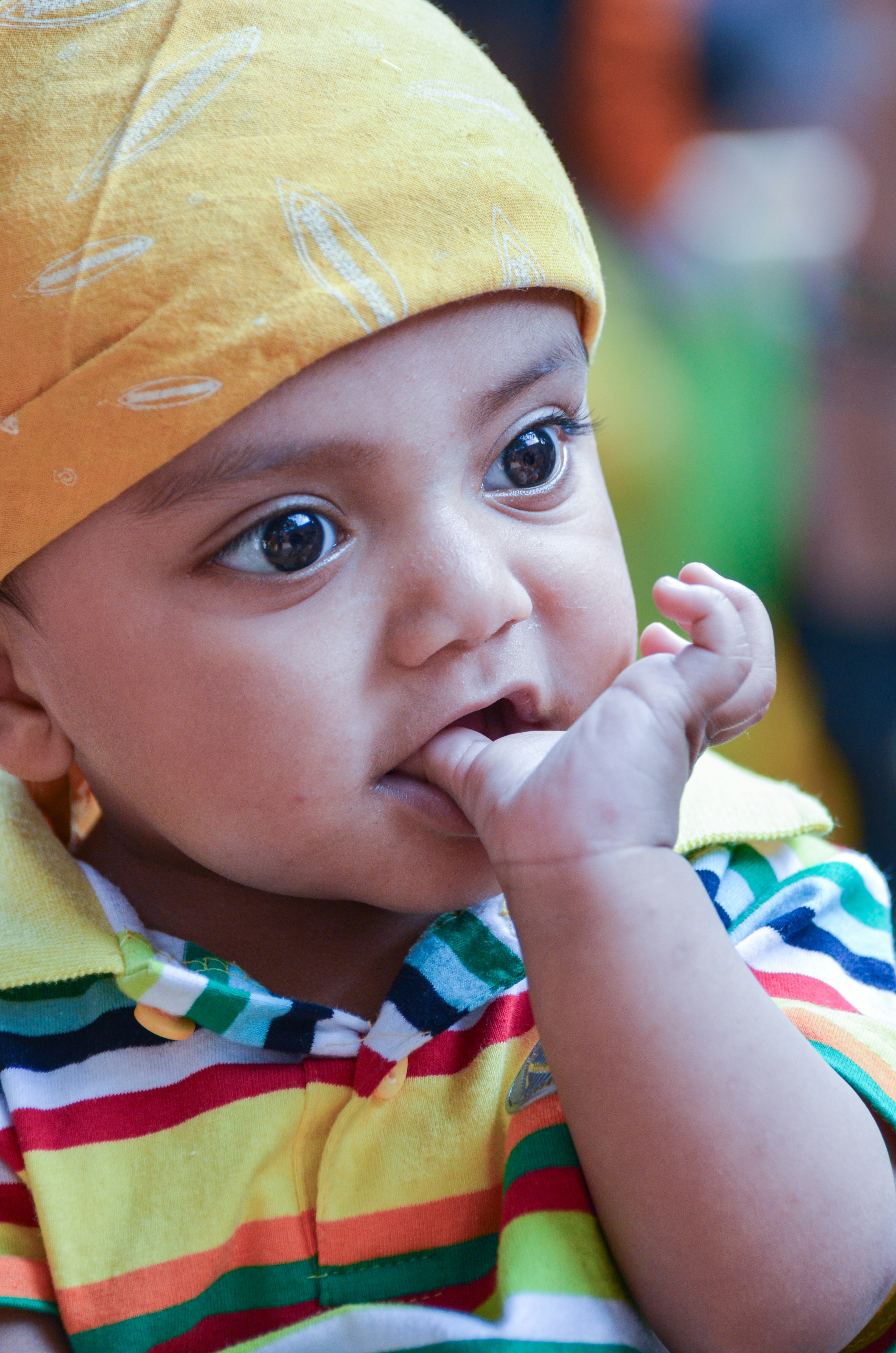 Close-up of a baby with wide, curious eyes wearing a yellow patterned headscarf and striped shirt; a candid moment highlighting innocence and wonder.