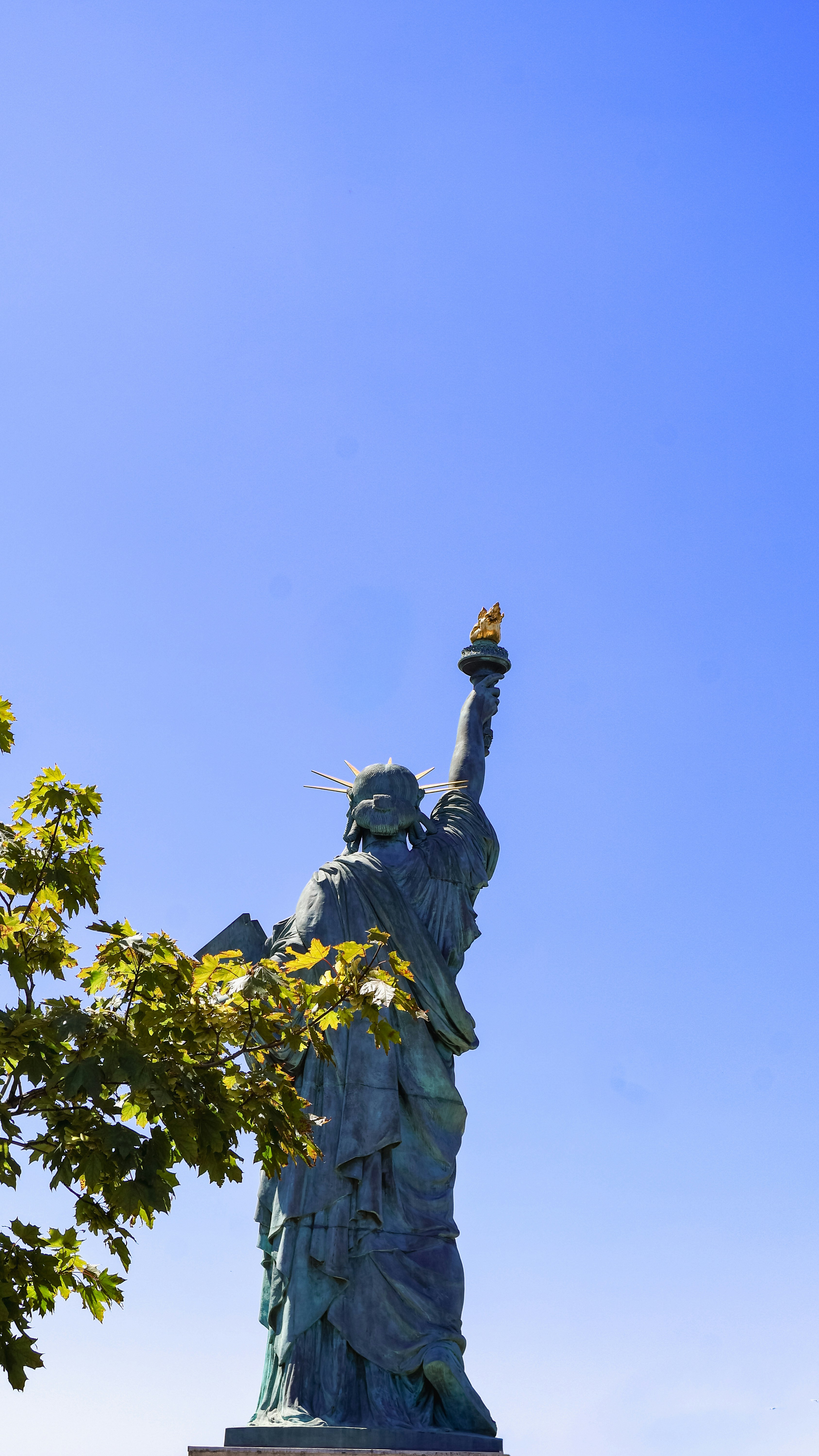 Bronze Statue of Liberty against a bright blue sky, with green leaves partially framing the lower left; a distant civic monument captured from ground level.