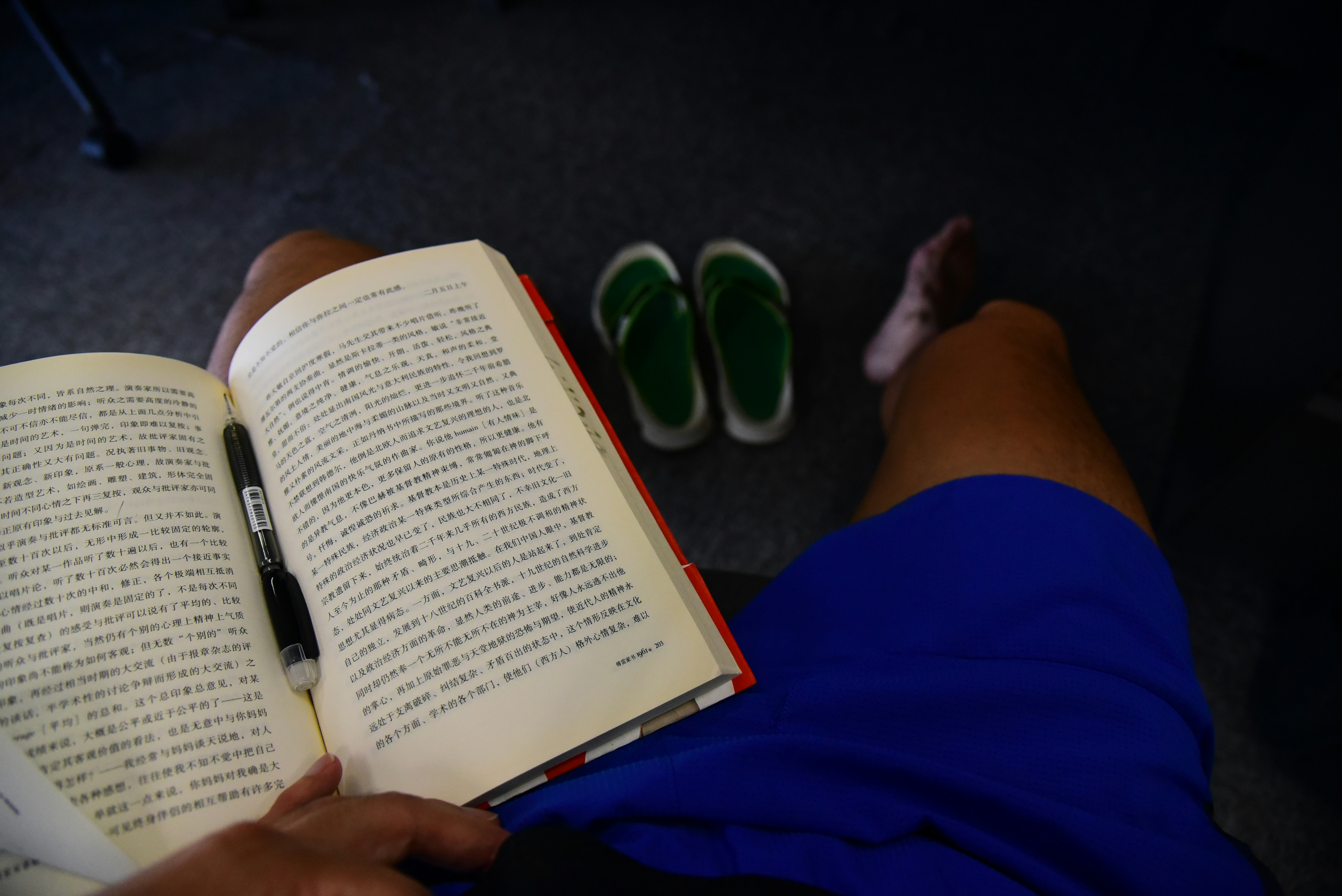 Open book held on a lap with a pen resting on a page; green shoes visible in the background under a blue garment, suggesting a casual reading moment.
