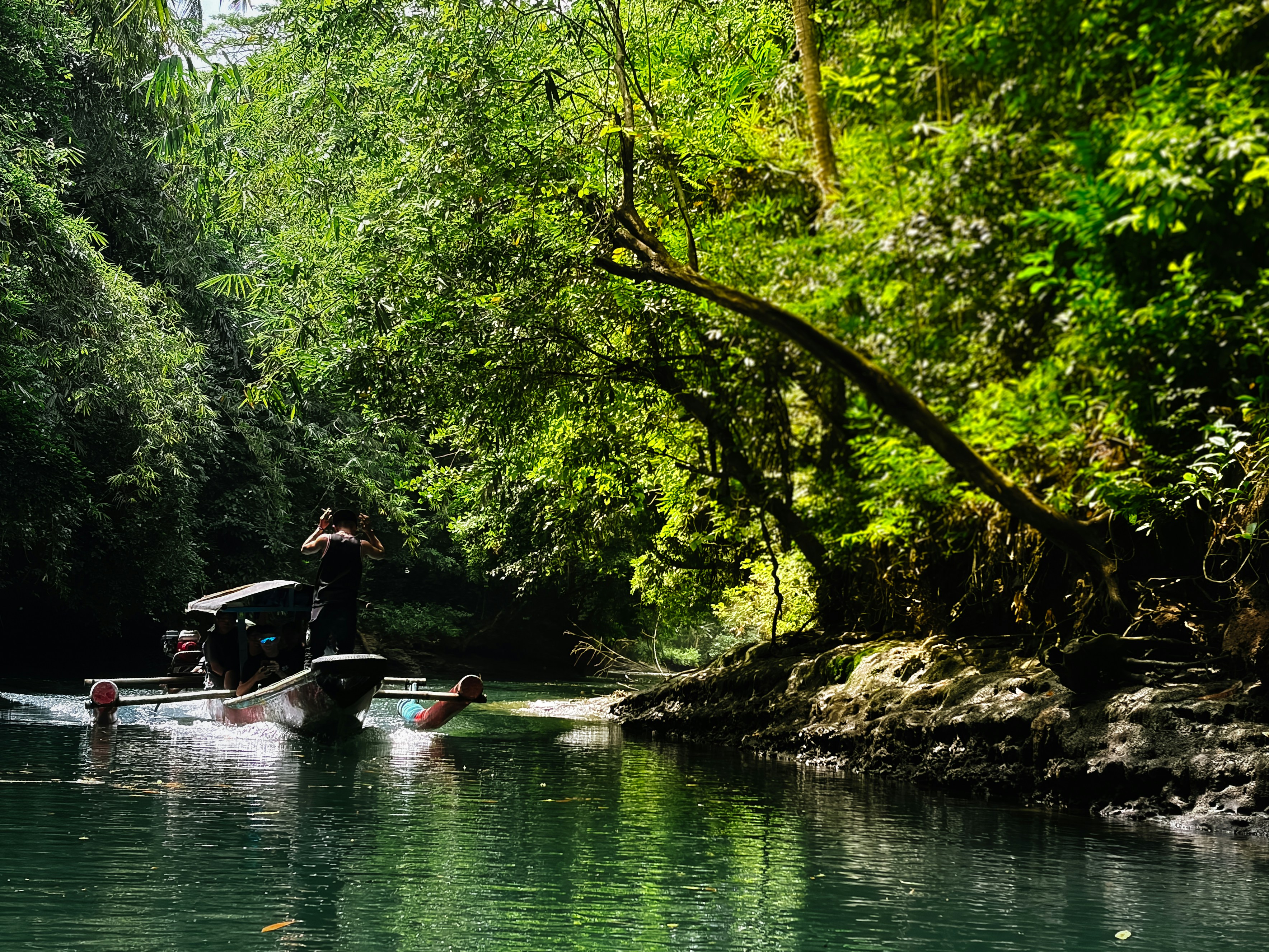 A small boat with people paddling along a calm river, shaded by lush green trees on both banks. The scene emphasizes the tranquil water and natural surroundings, suggesting a quiet excursion in a forested gorge.