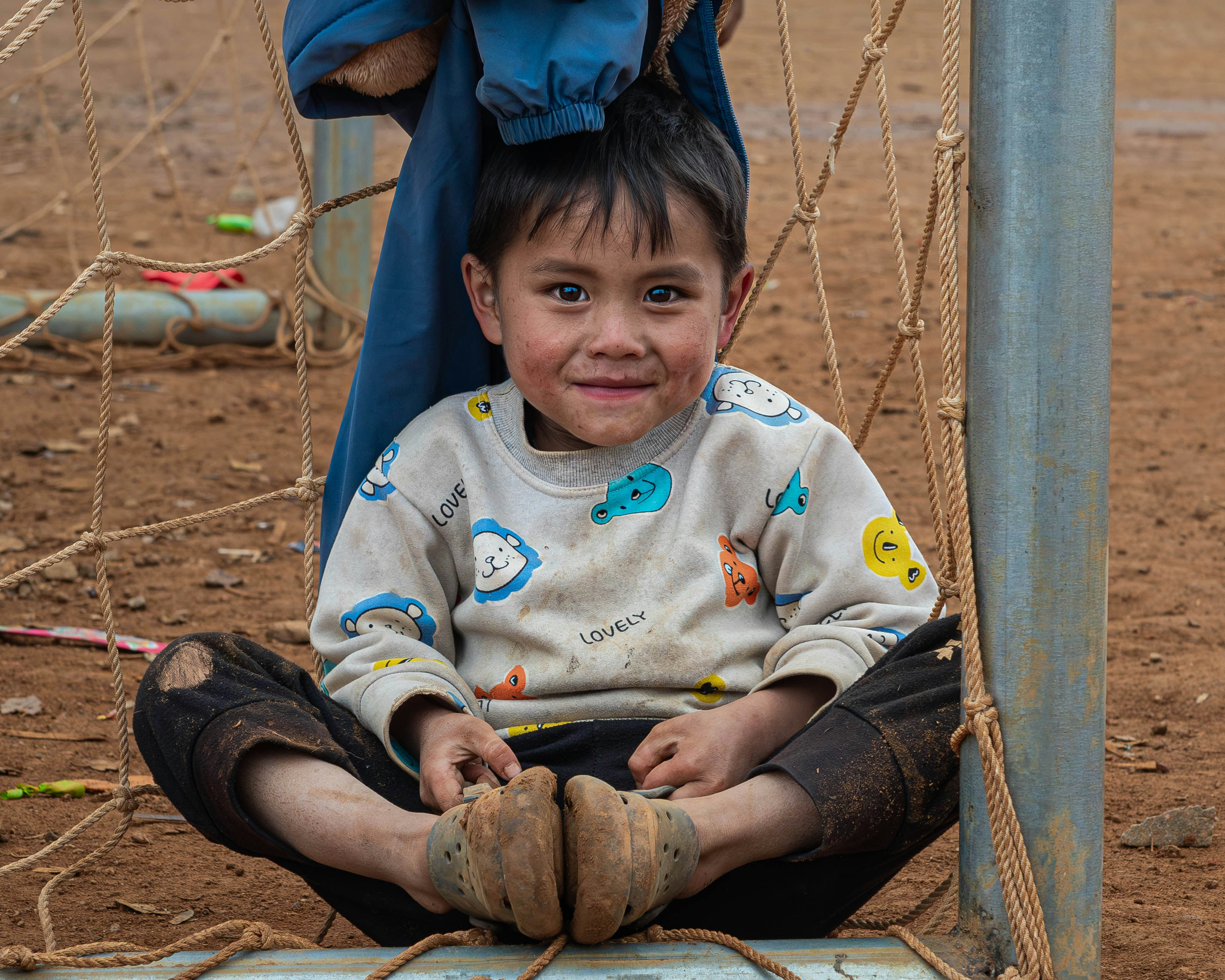 A child in a patterned sweatshirt sitting on dirt inside a rope climbing frame