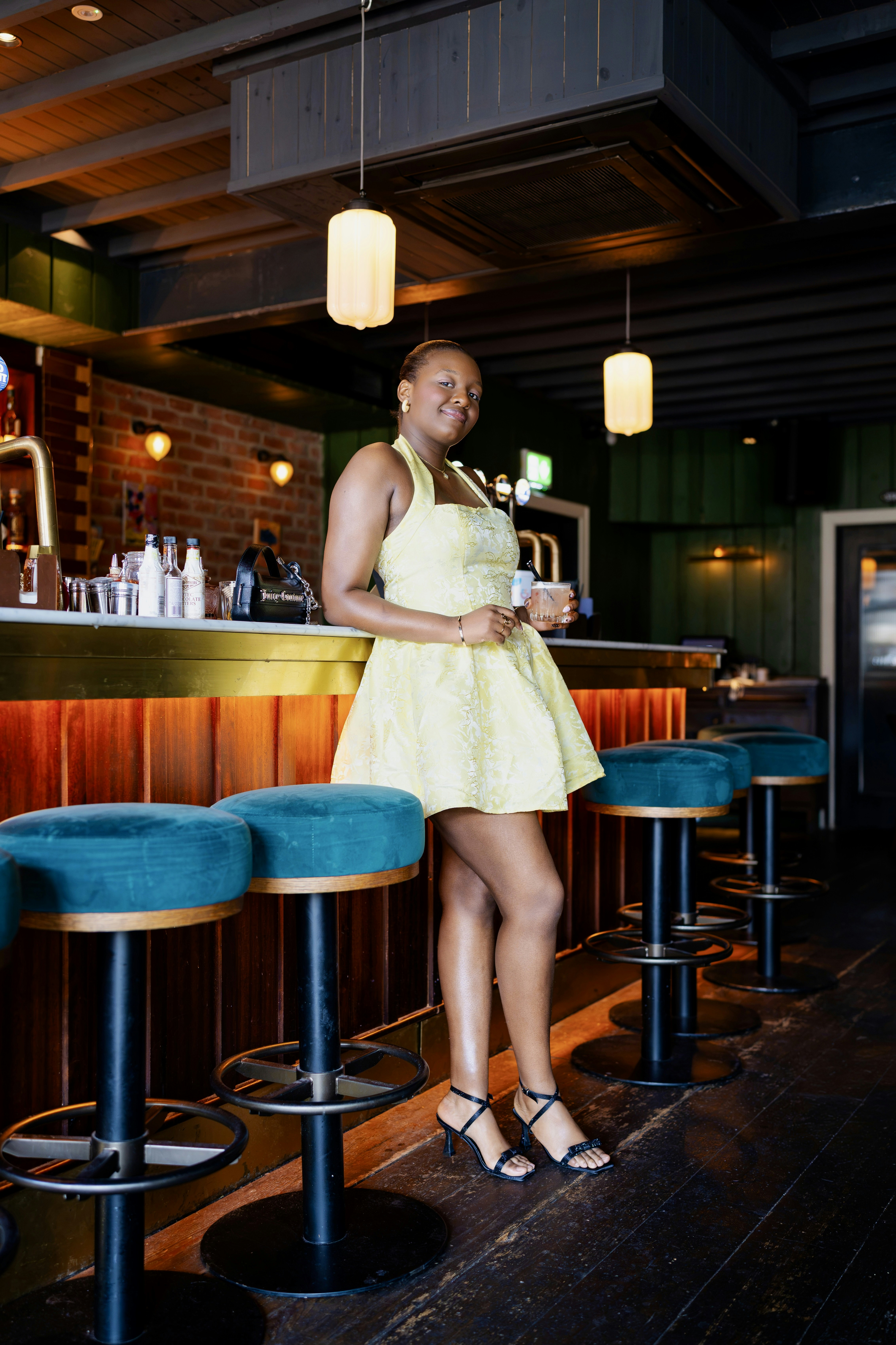 A stylish woman in a pale yellow dress leans against a warm-lit bar in a contemporary lounge; the scene highlights the bar stools, brick wall, and ambient pendant lighting.