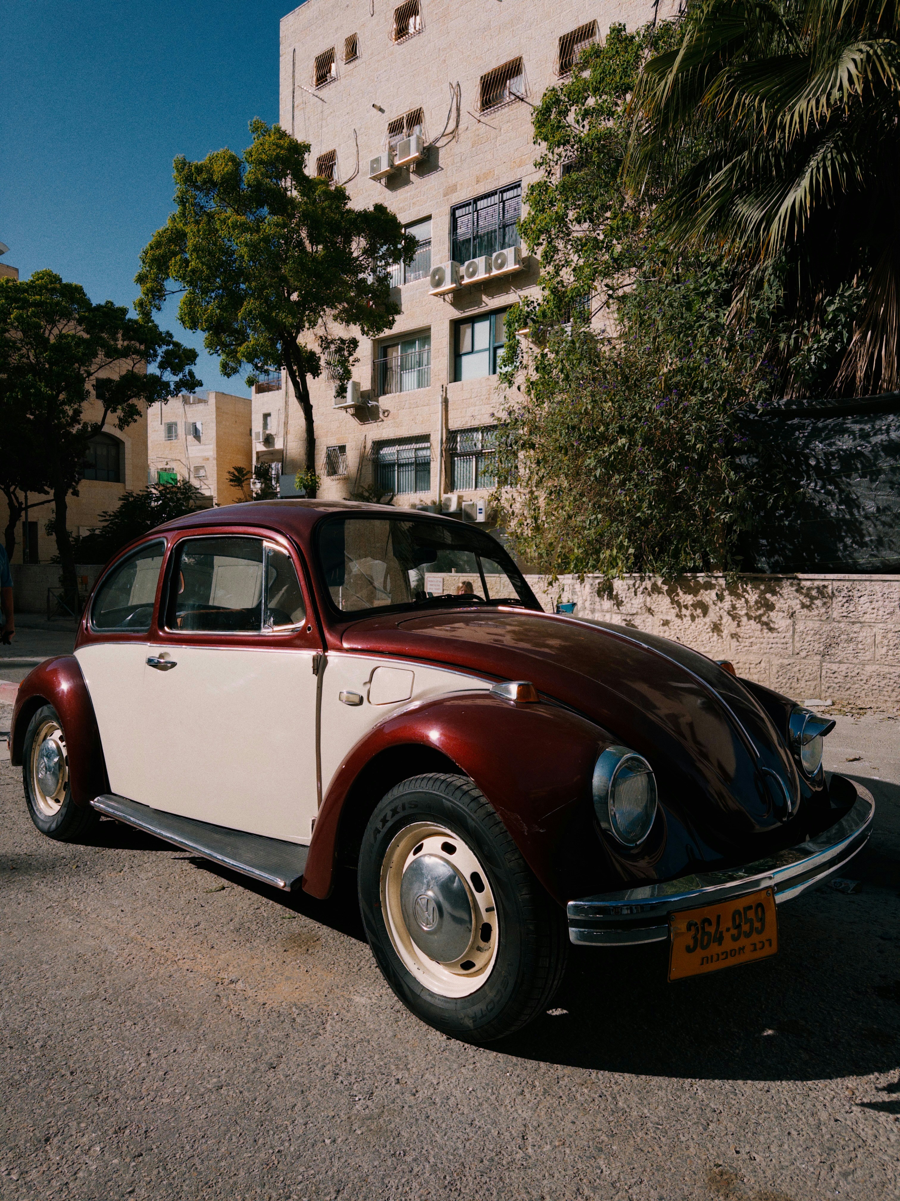 A burgundy and cream vintage Volkswagen Beetle parked on a sunlit street, with a mid-century building and lush trees in the background. The shot emphasizes the car's two-tone body and classic curves.