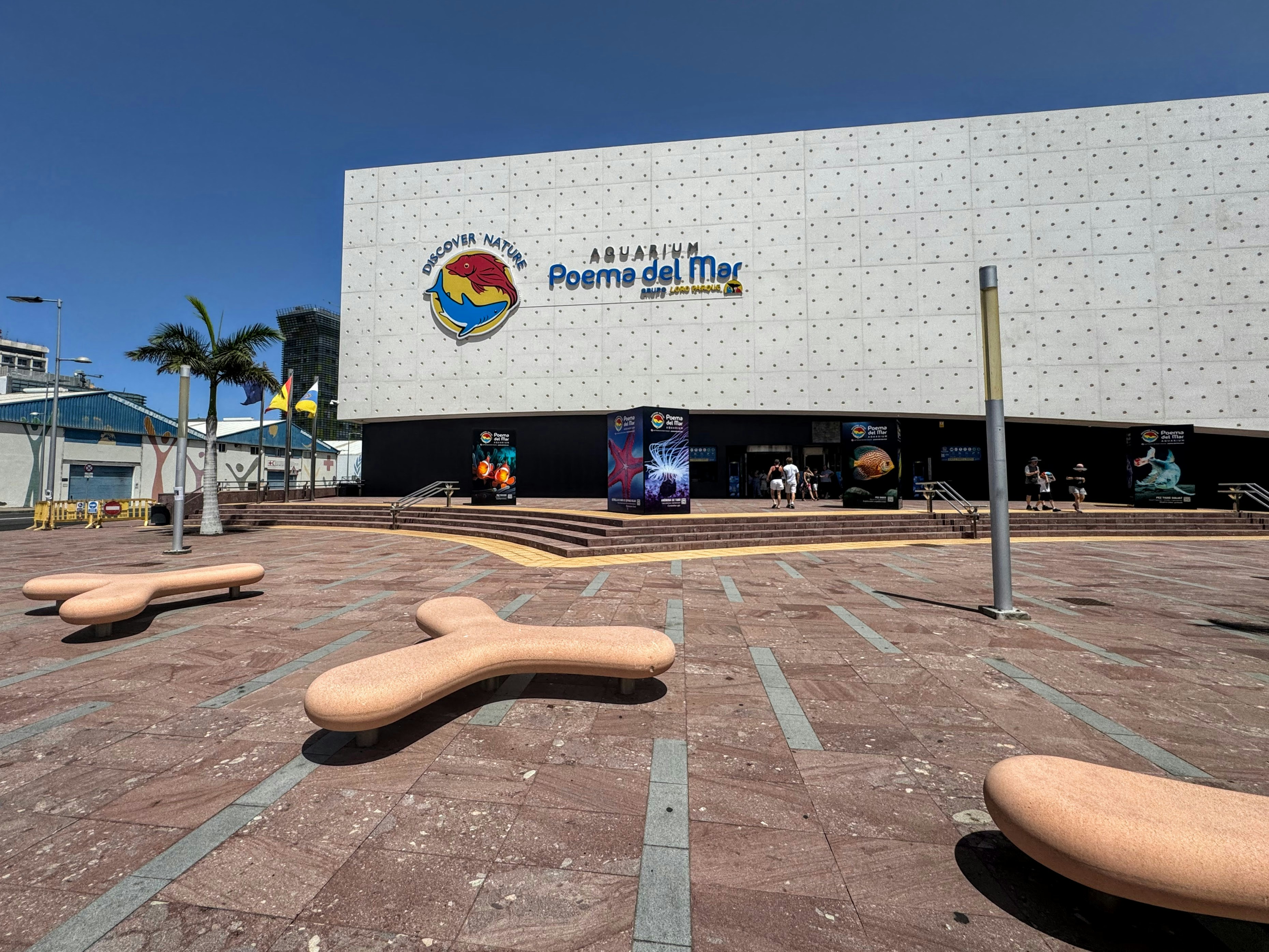 Wide-angle view of the Poema del Mar building with a patterned white facade, palm trees, and sculptural benches in a brick plaza.