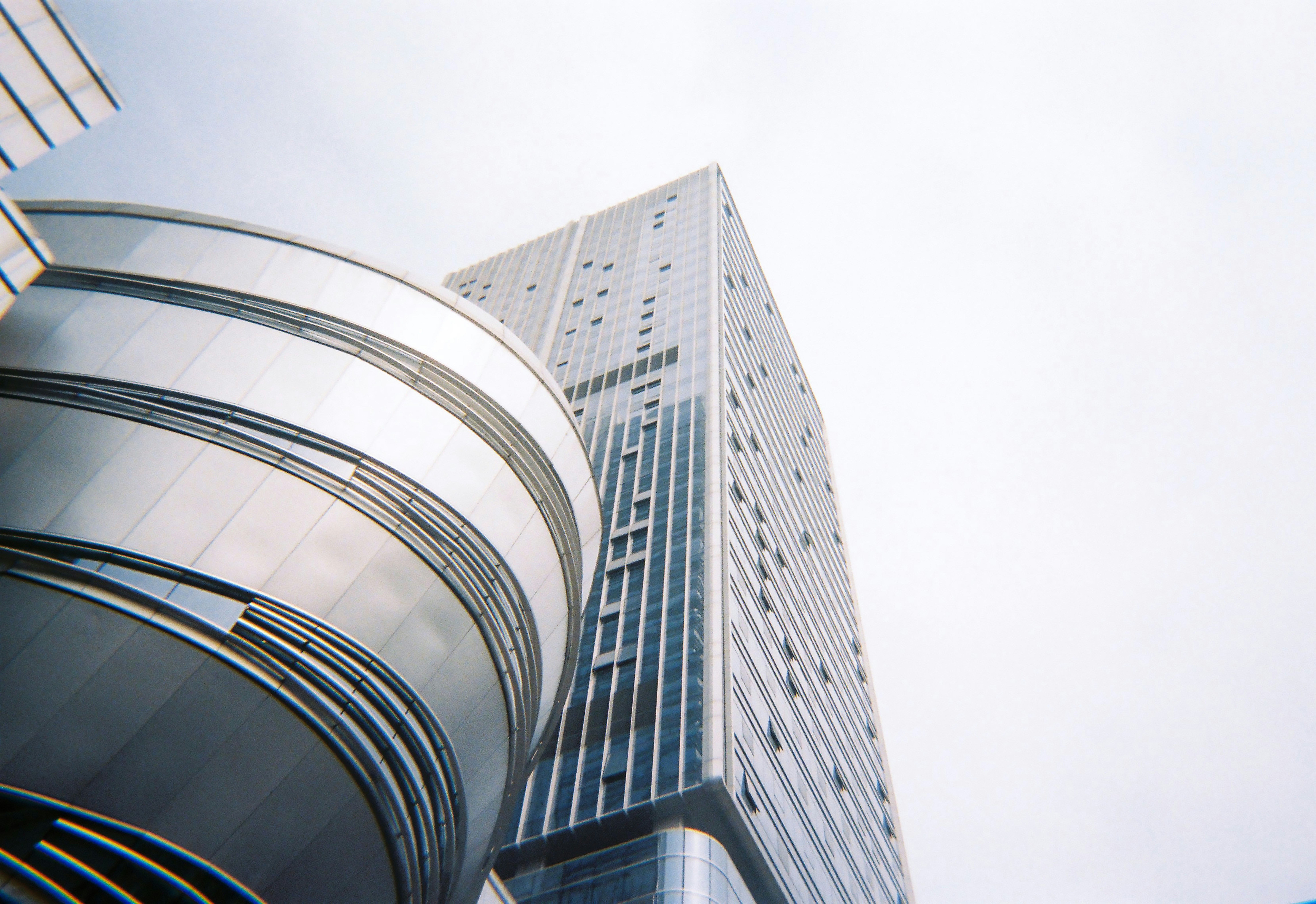 Modern architectural composition with curved metallic structure in foreground and tall glass tower reaching the pale sky.