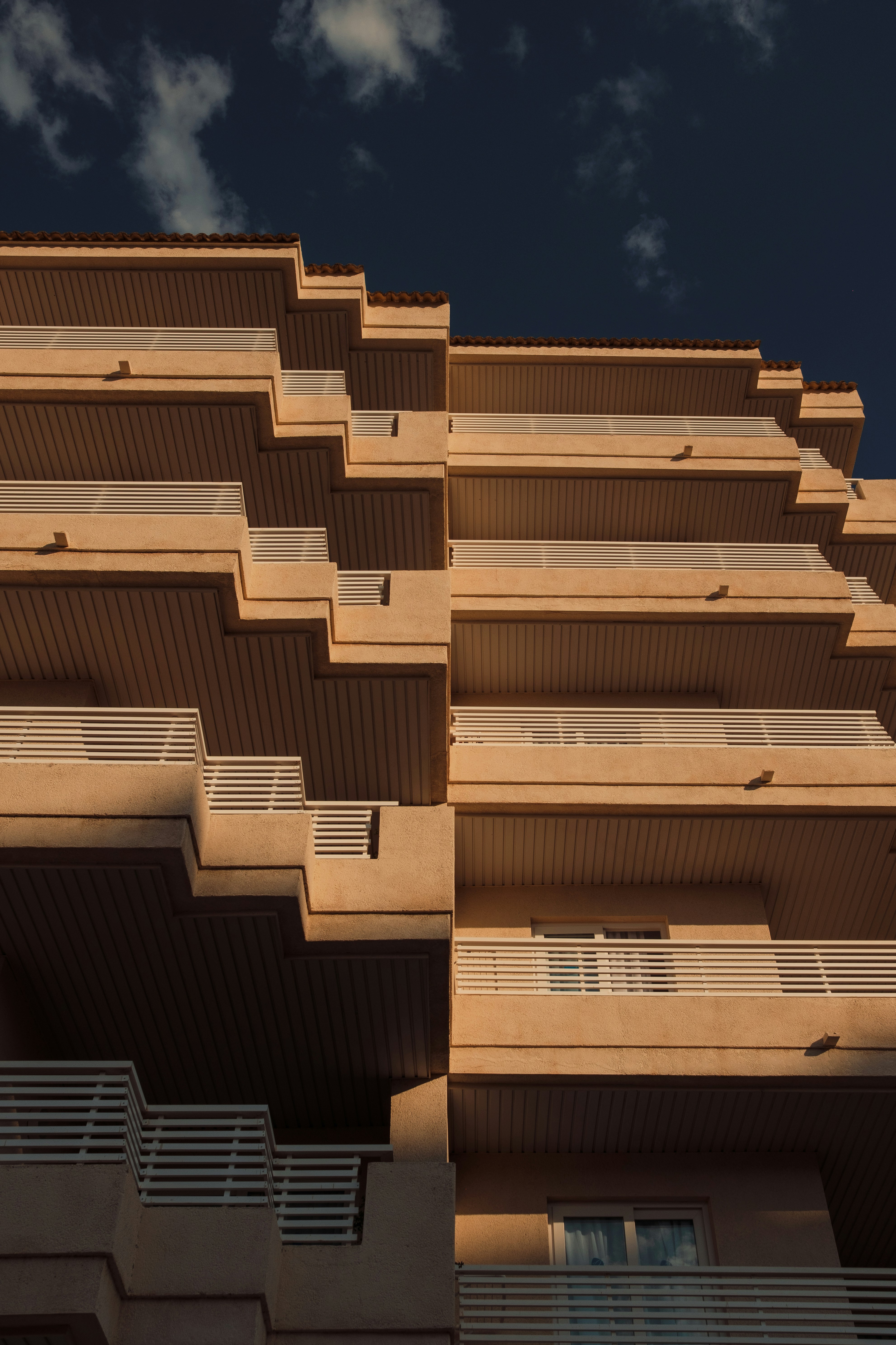 A low-angle graphic photograph of a warm, peach-toned resort building in Mallorca, highlighting the stepped geometric patterns of stacked balconies and dramatic light-and-shadow contrast against a deep blue sky.