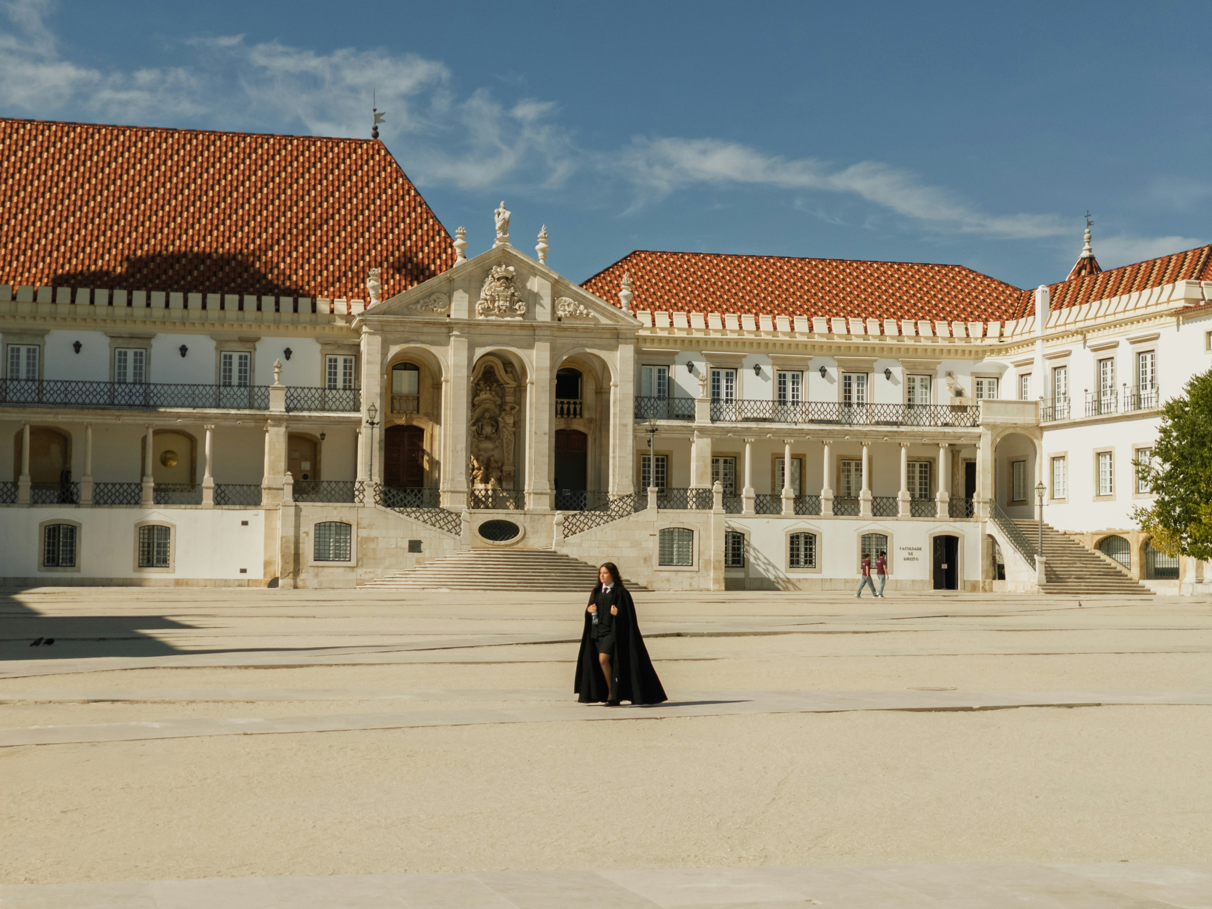 A lone figure in dark robes stands in a vast open square before a historic building with a red-tiled roof and ornate facade.