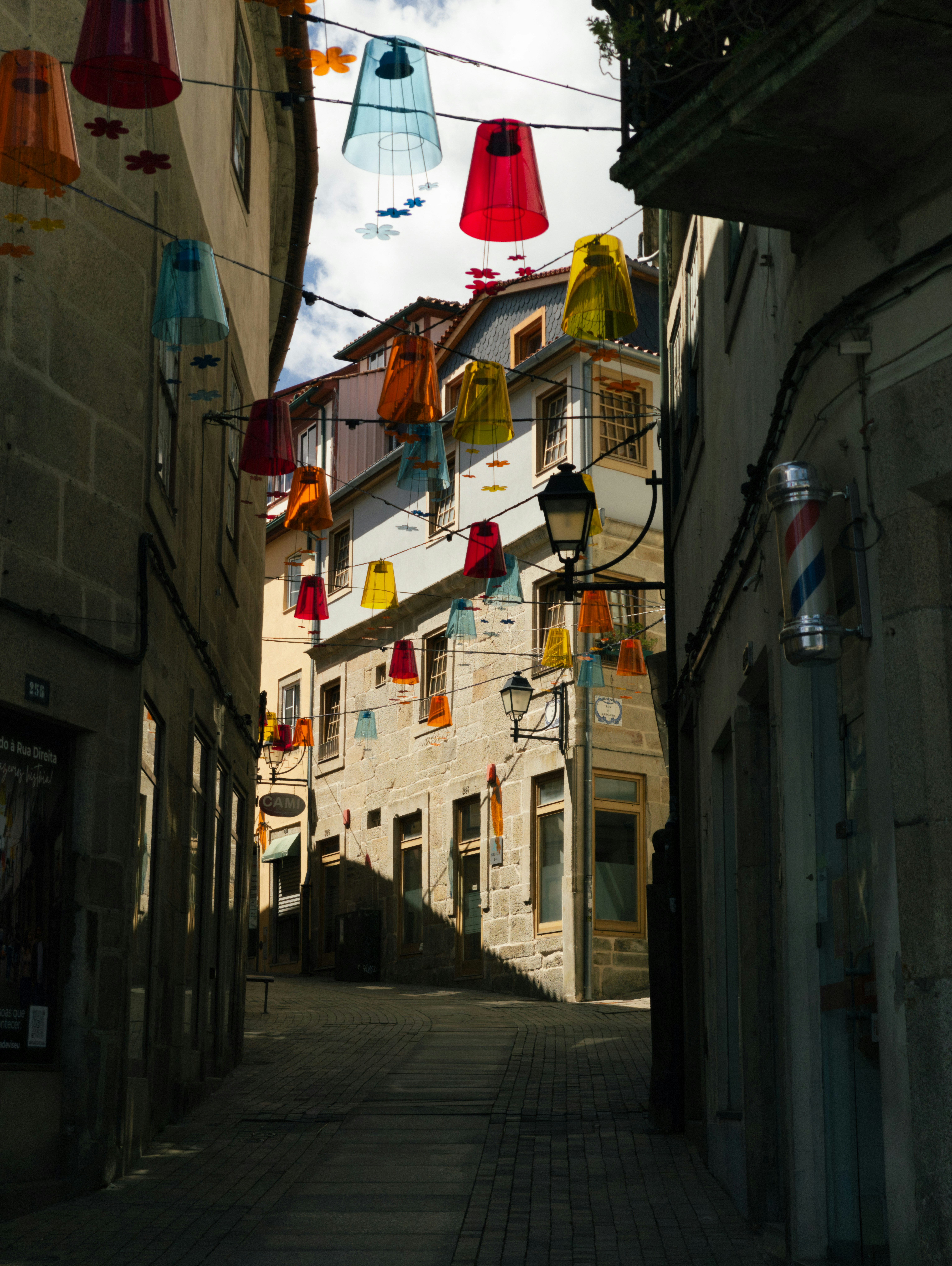 Sunlit narrow alley lined with stone buildings, colorful lanterns strung overhead casting a warm glow.