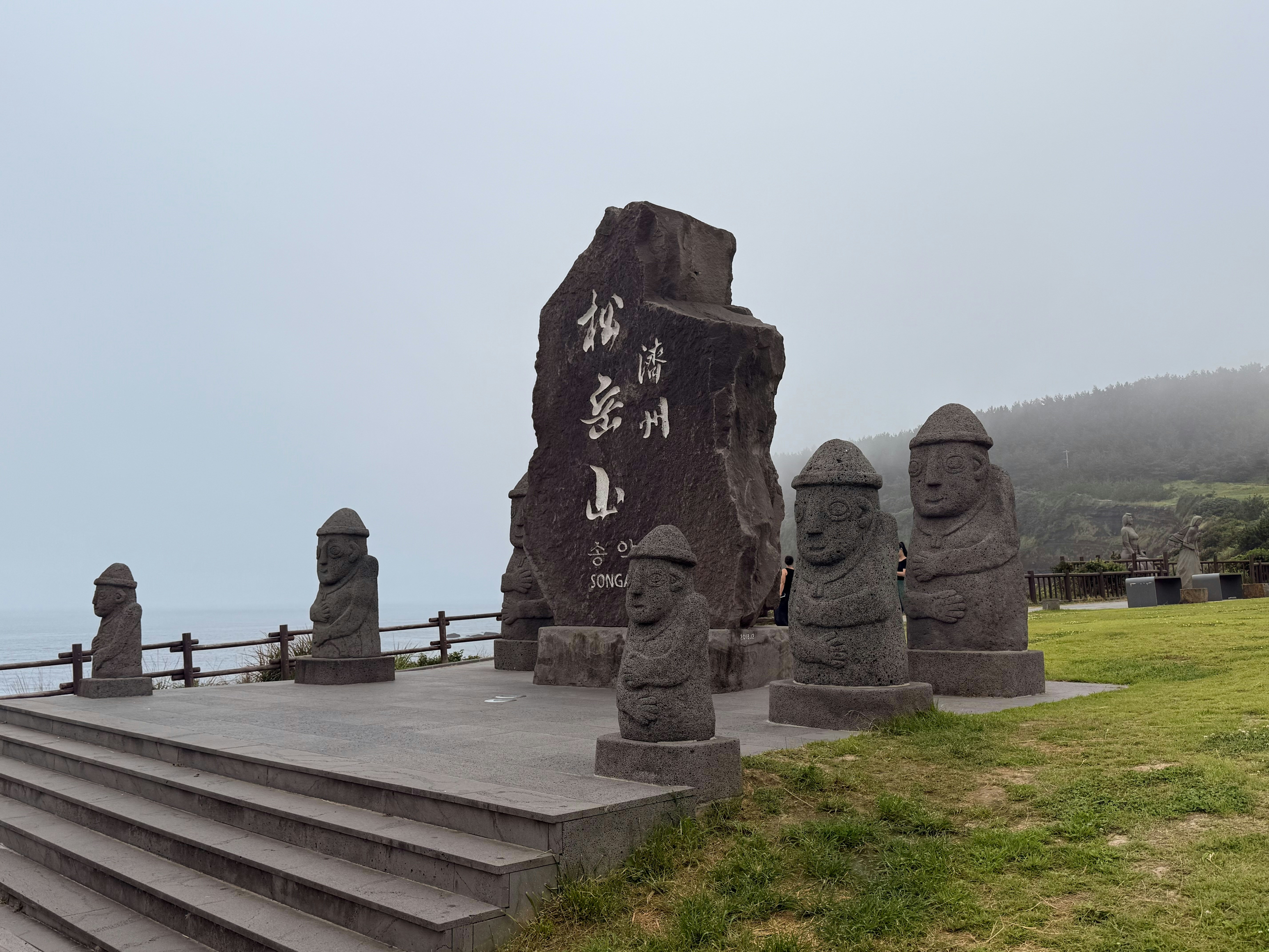 Row of carved stone statues along a terrace by the sea cliff, with a tall engraved rock and distant hillside in a foggy scene.
