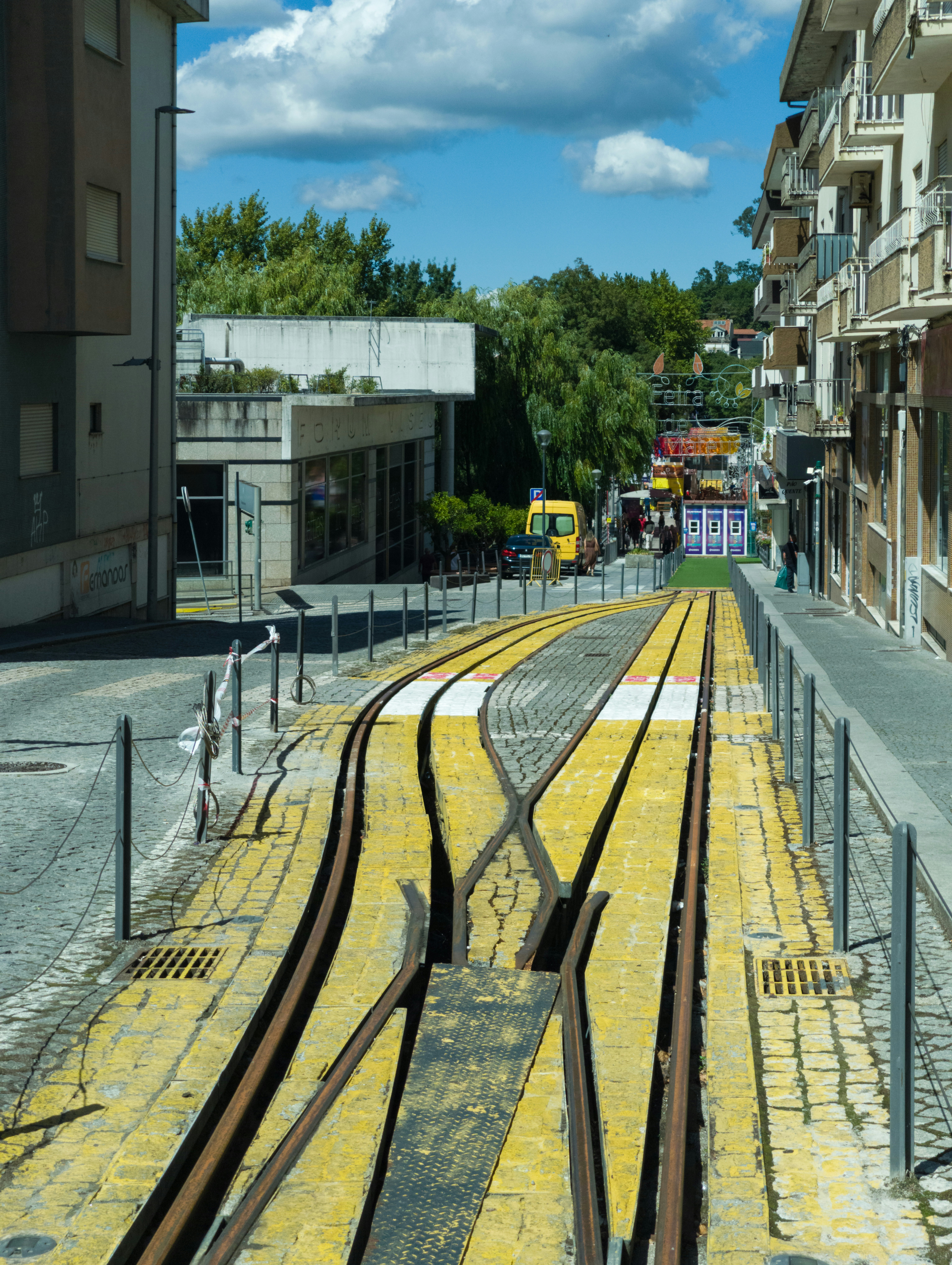 Yellow tram rails arc through a sunlit urban street, flanked by buildings and a distant van. A concrete platform and pedestrians hint at a public transit corridor in an intersection-filled city.