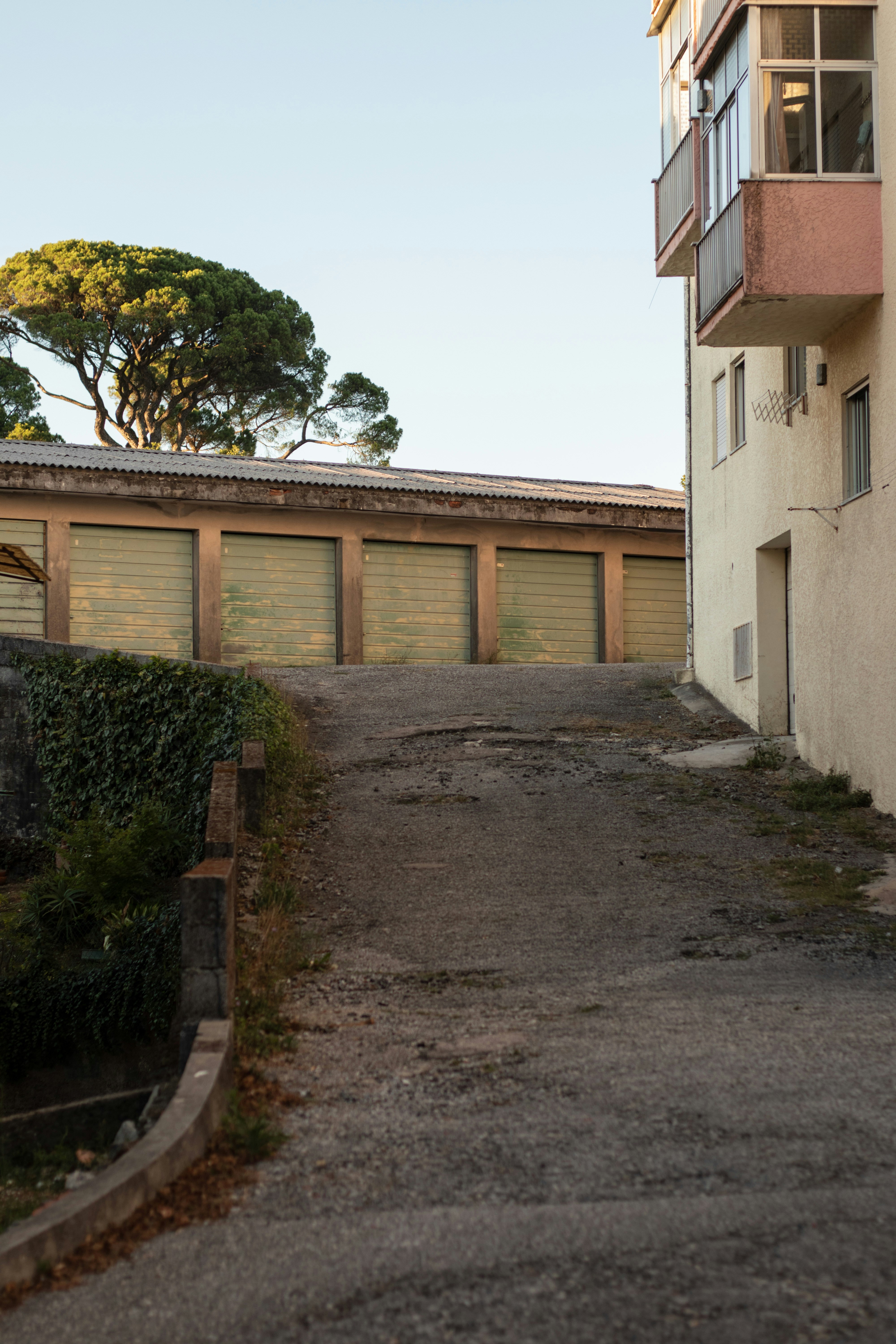 Sunlit path beside a building slopes upward toward a row of closed garage doors, with a tree peeking over the roof. A quiet, urban incline scene capturing texture and light in a candid street environment.