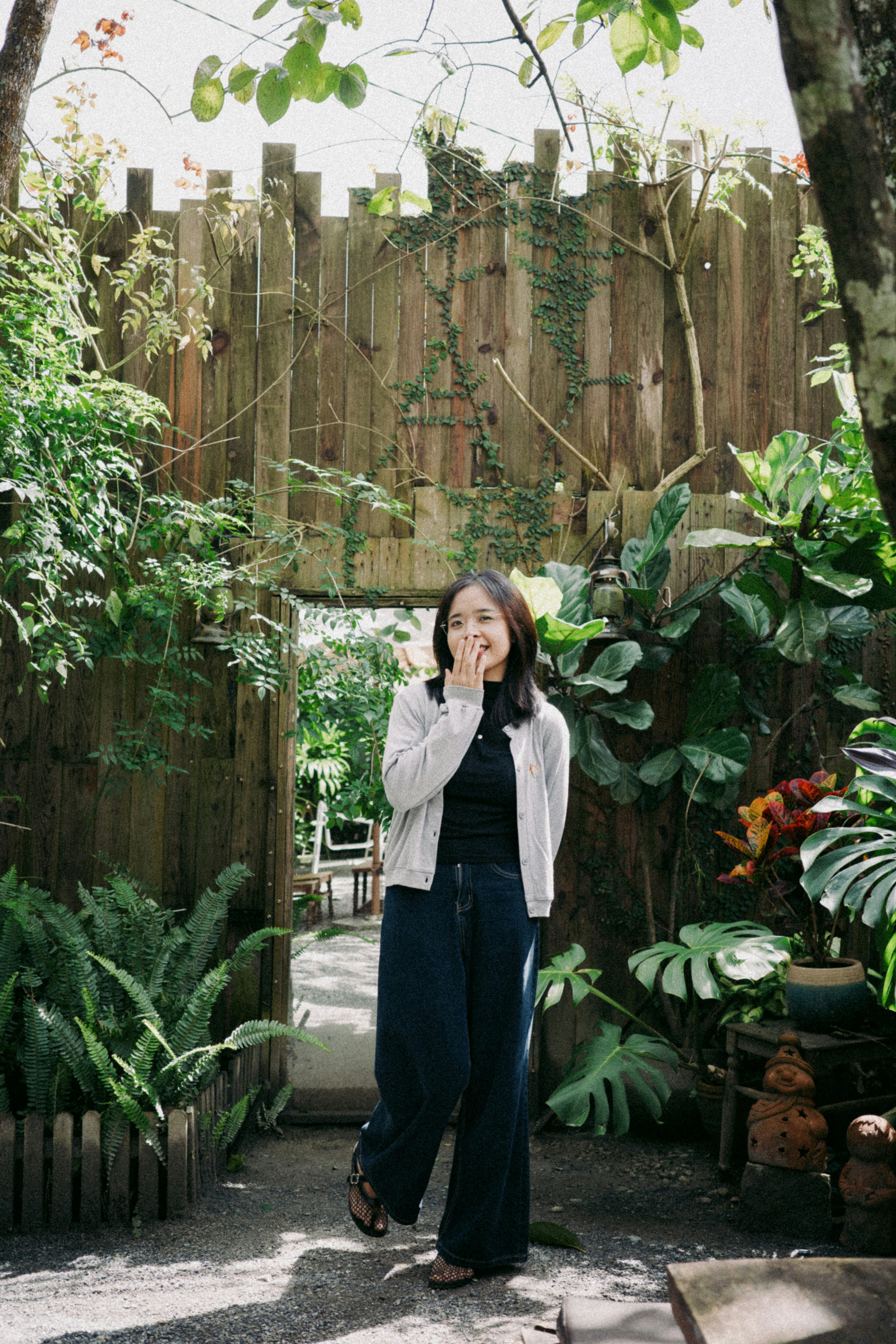 A young woman stands in a sunlit greenhouse doorway, fingers near her lips, surrounded by lush plants and a wooden fence. The scene feels intimate and serene, with natural textures framing the subject.