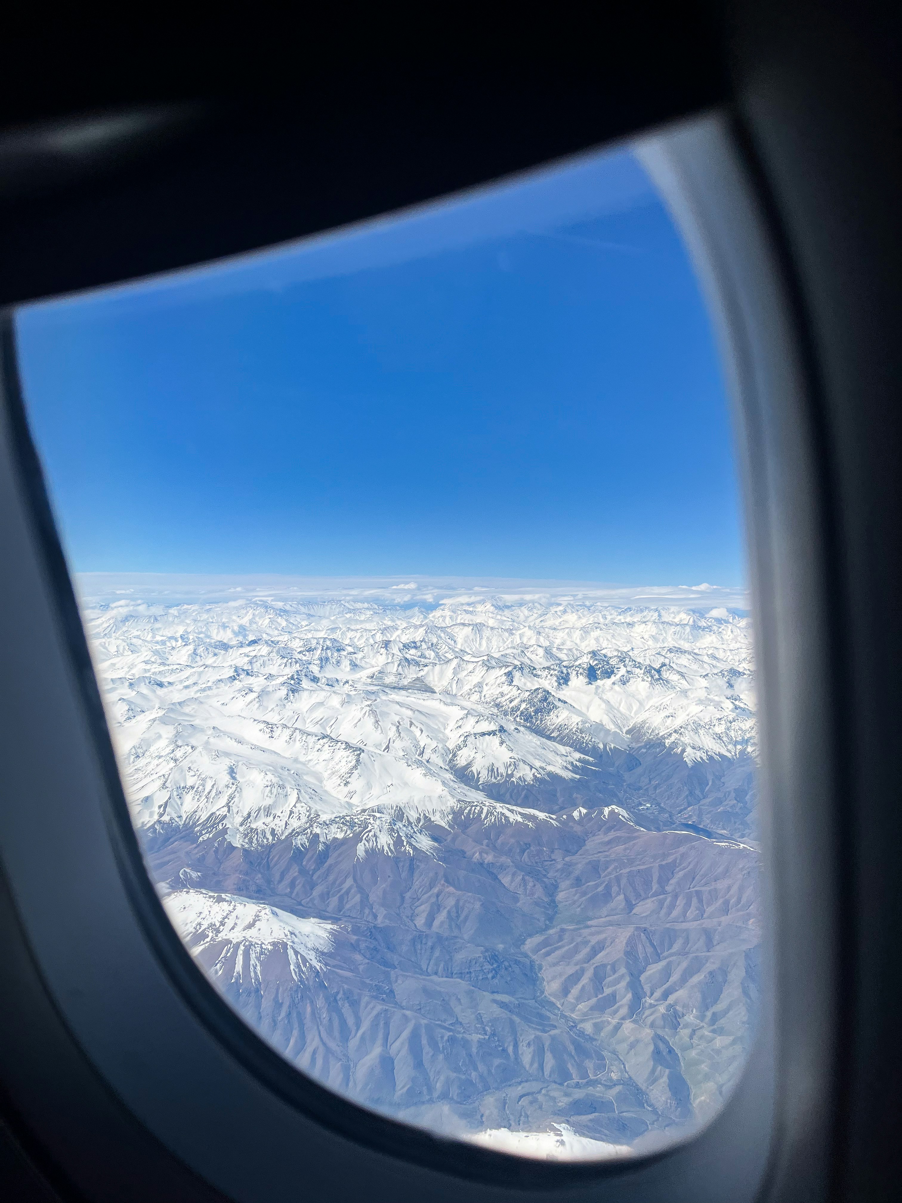 Snowy mountain range seen through an aircraft window, with the blue sky stretching above and the window frame providing a natural vignette.