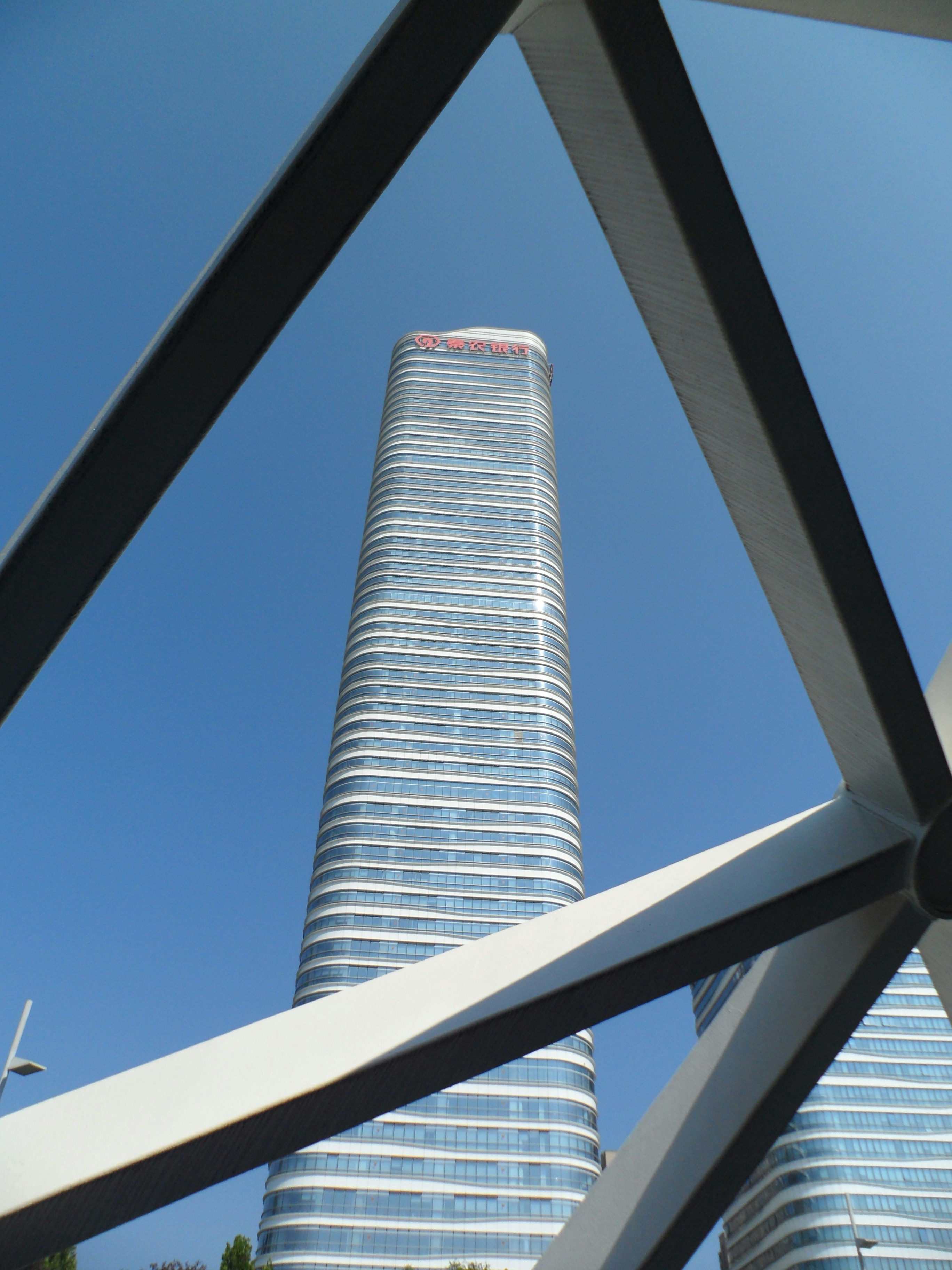 Tall glass-clad tower piercing a clear blue sky, framed by bold steel trusses in the foreground. A modern architectural moment captured through a lattice backdrop.