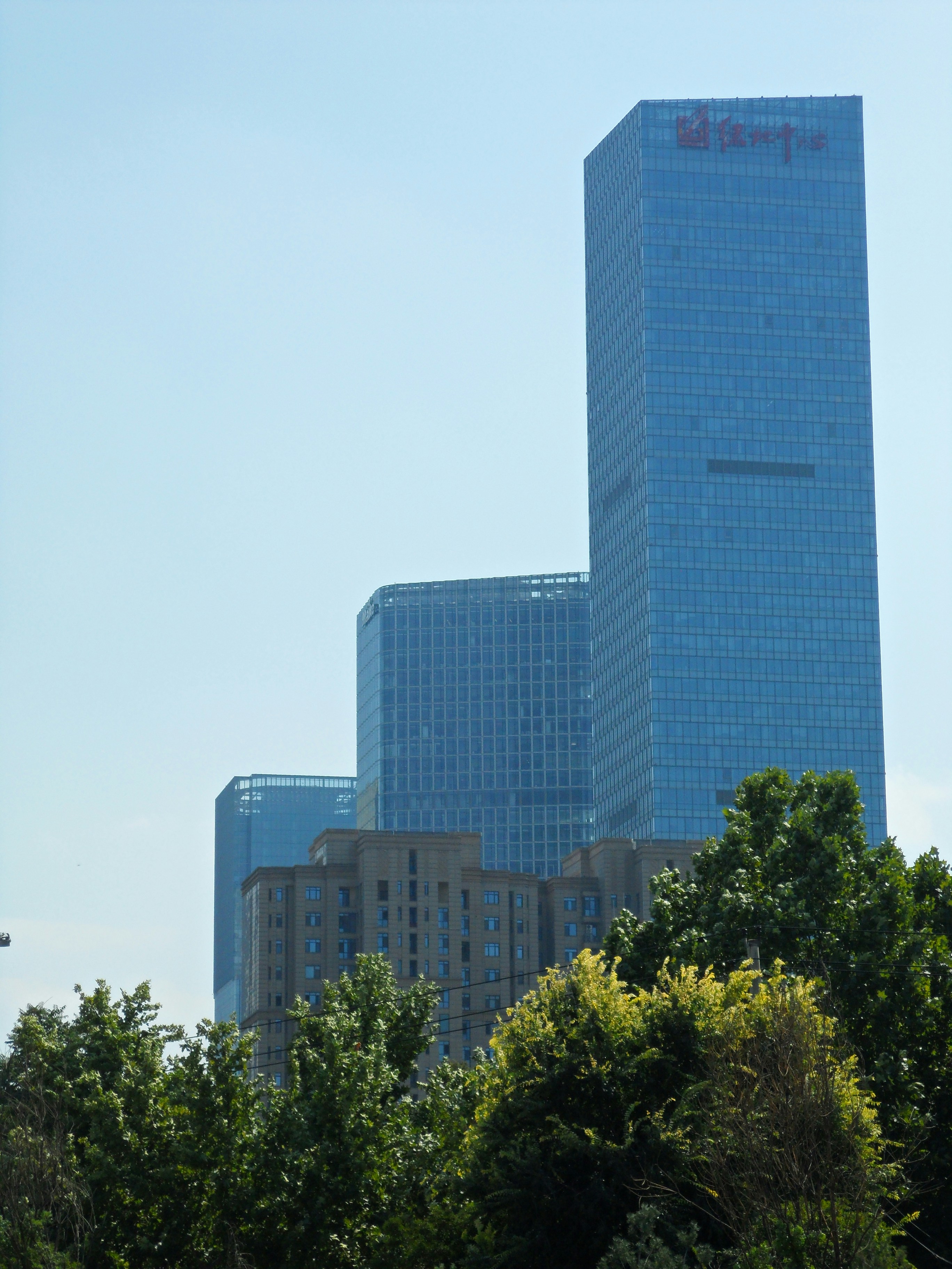 Modern blue glass skyscrapers rising behind tan apartment buildings and green trees