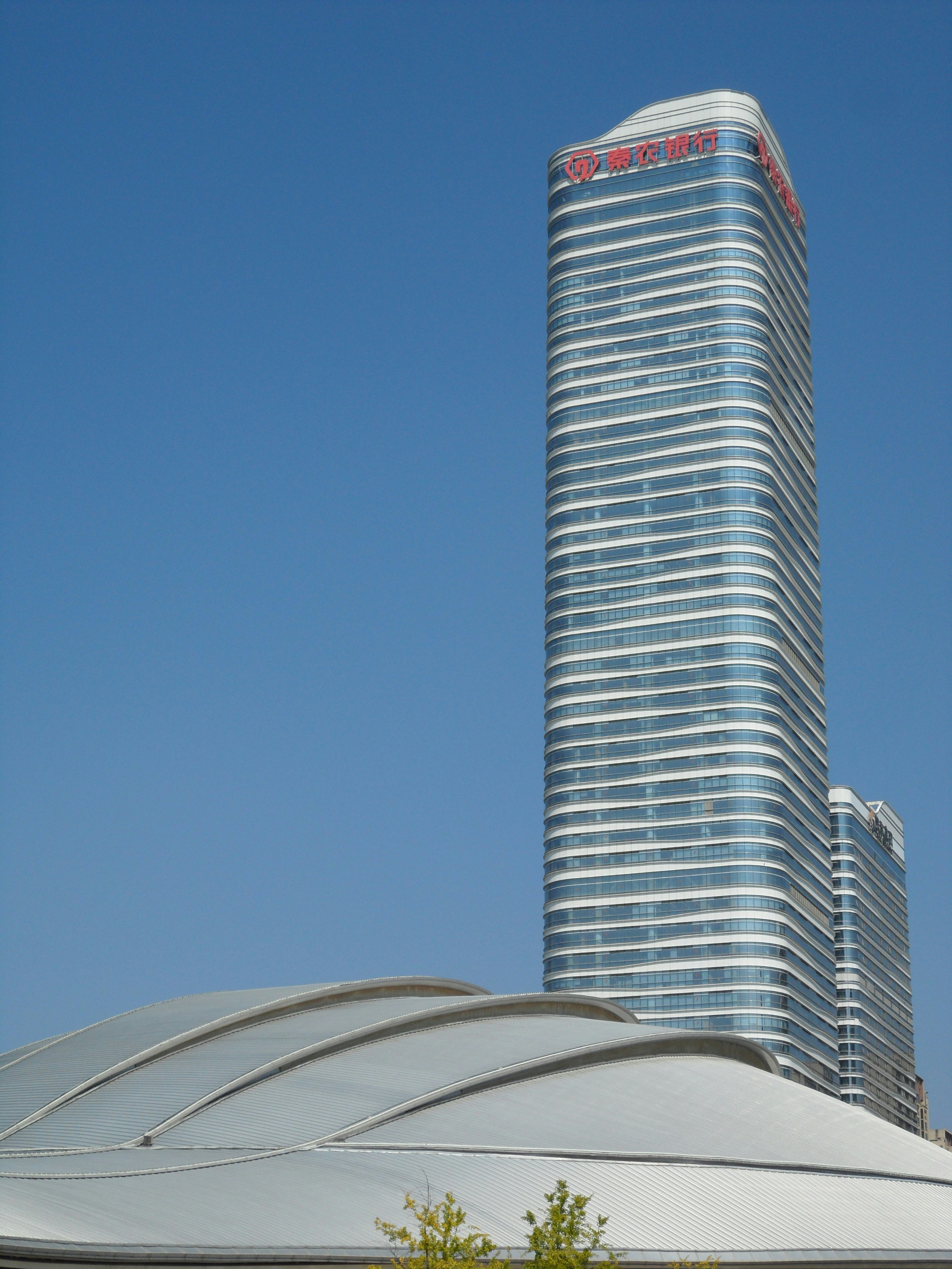 A sleek, curvilinear skyscraper rises against a clear blue sky beside a modern curved-roof complex.