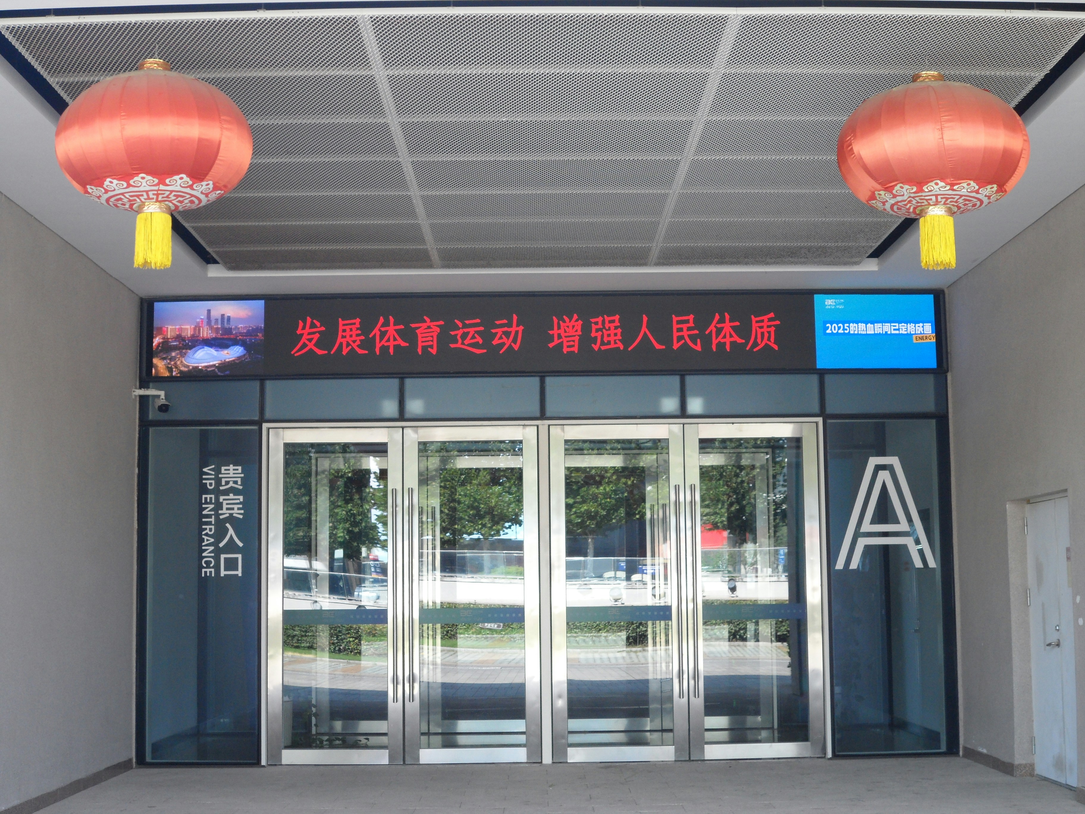 Glass double-doors framed by a modern facade with red LED banner and two decorative red lanterns above, suggesting a public or cultural facility.