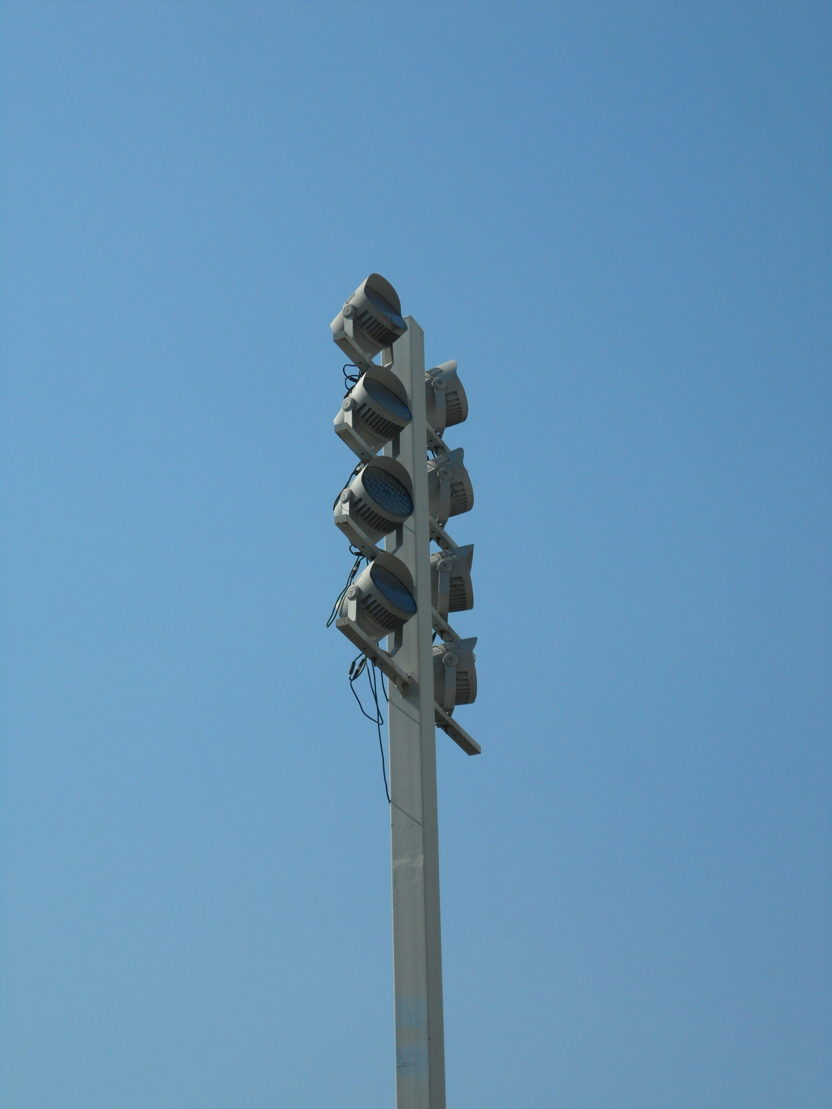 Tall stadium floodlight tower against a cloudless blue sky, captured with minimal surrounding context to emphasize industrial structure. This is a photographic study of form and engineering rather than activity.