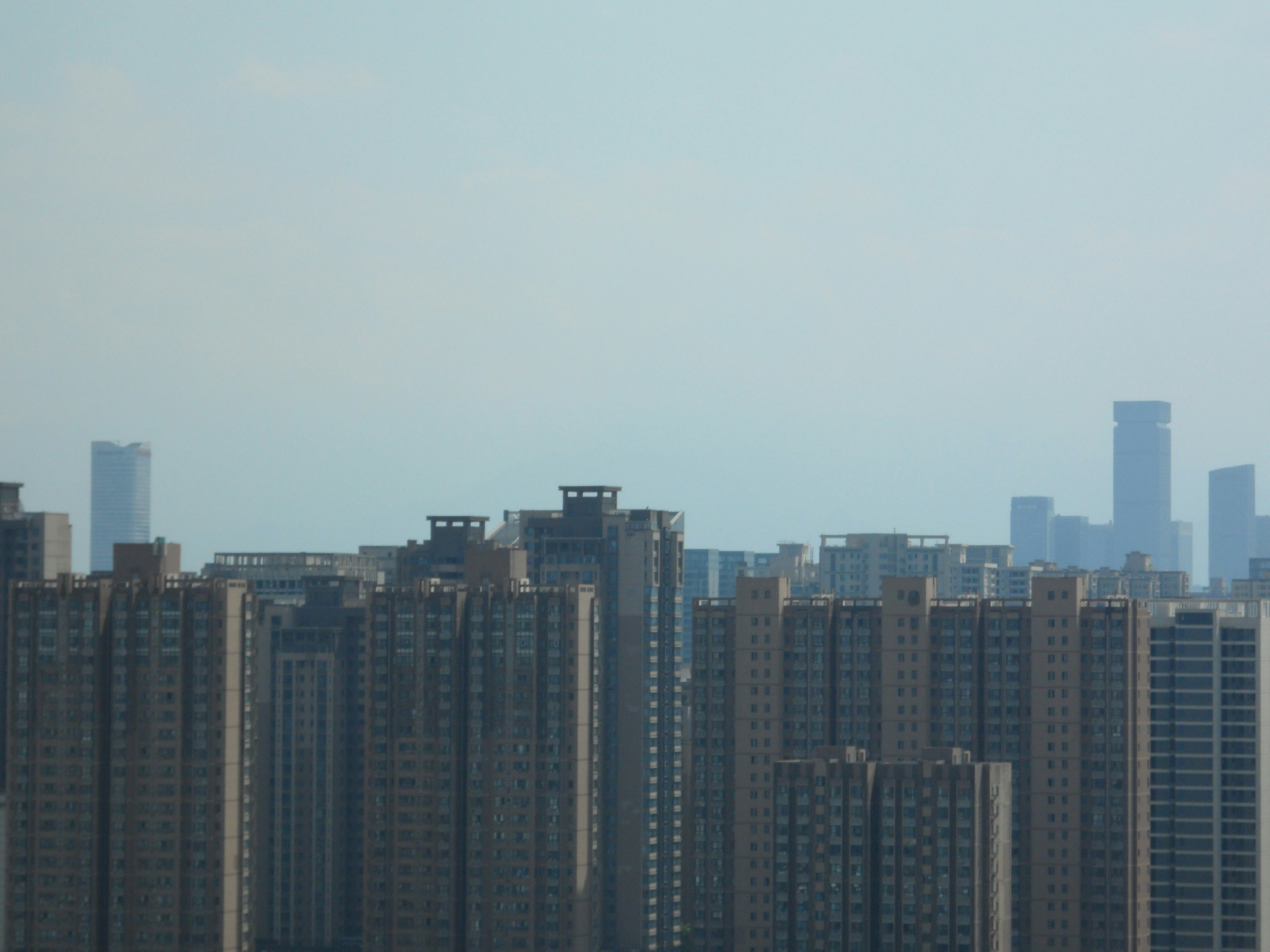 Dense residential towers rise in a uniform grid beneath a pale sky, creating a cityscape silhouette with distant high-rises on the horizon.