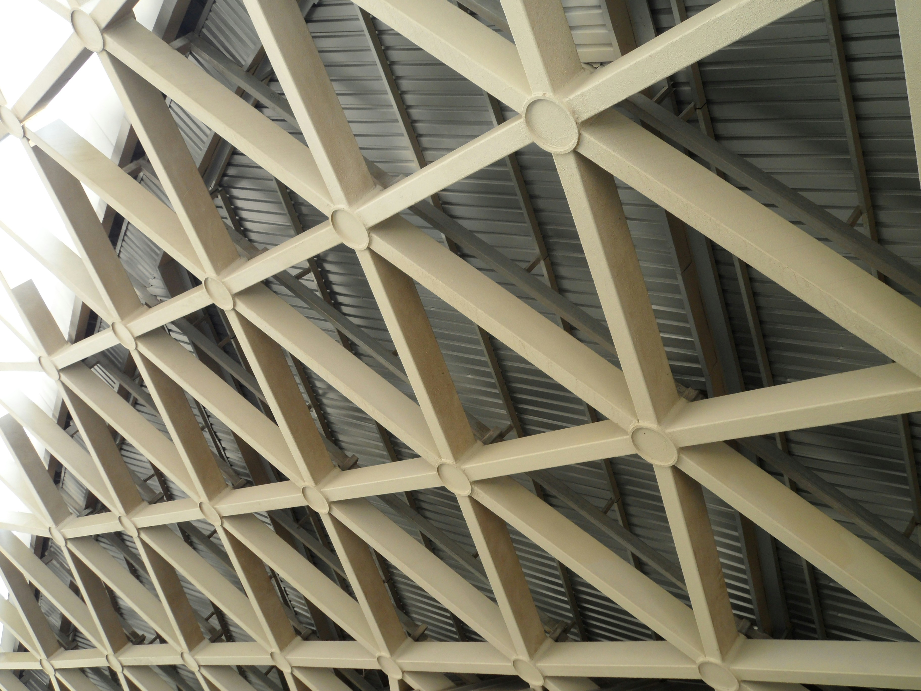 Architectural lattice of beige steel beams creates a repeating geometric pattern across the frame, with soft light filtering in from the left. The perspective emphasizes depth and structure rather than a human subject.