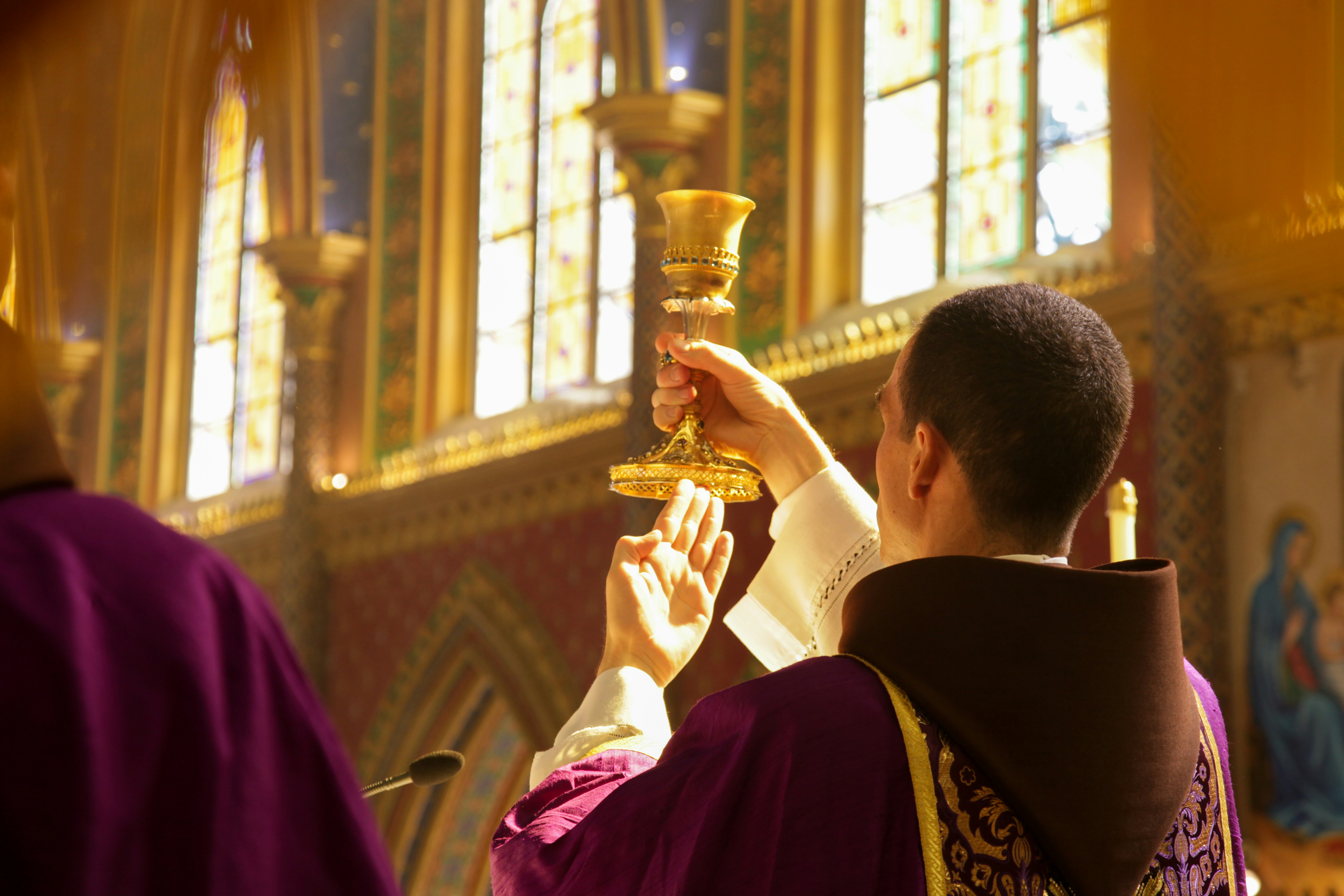 A priest in purple vestments raises a ceremonial chalice during a gilded church service, bathed in warm window light.