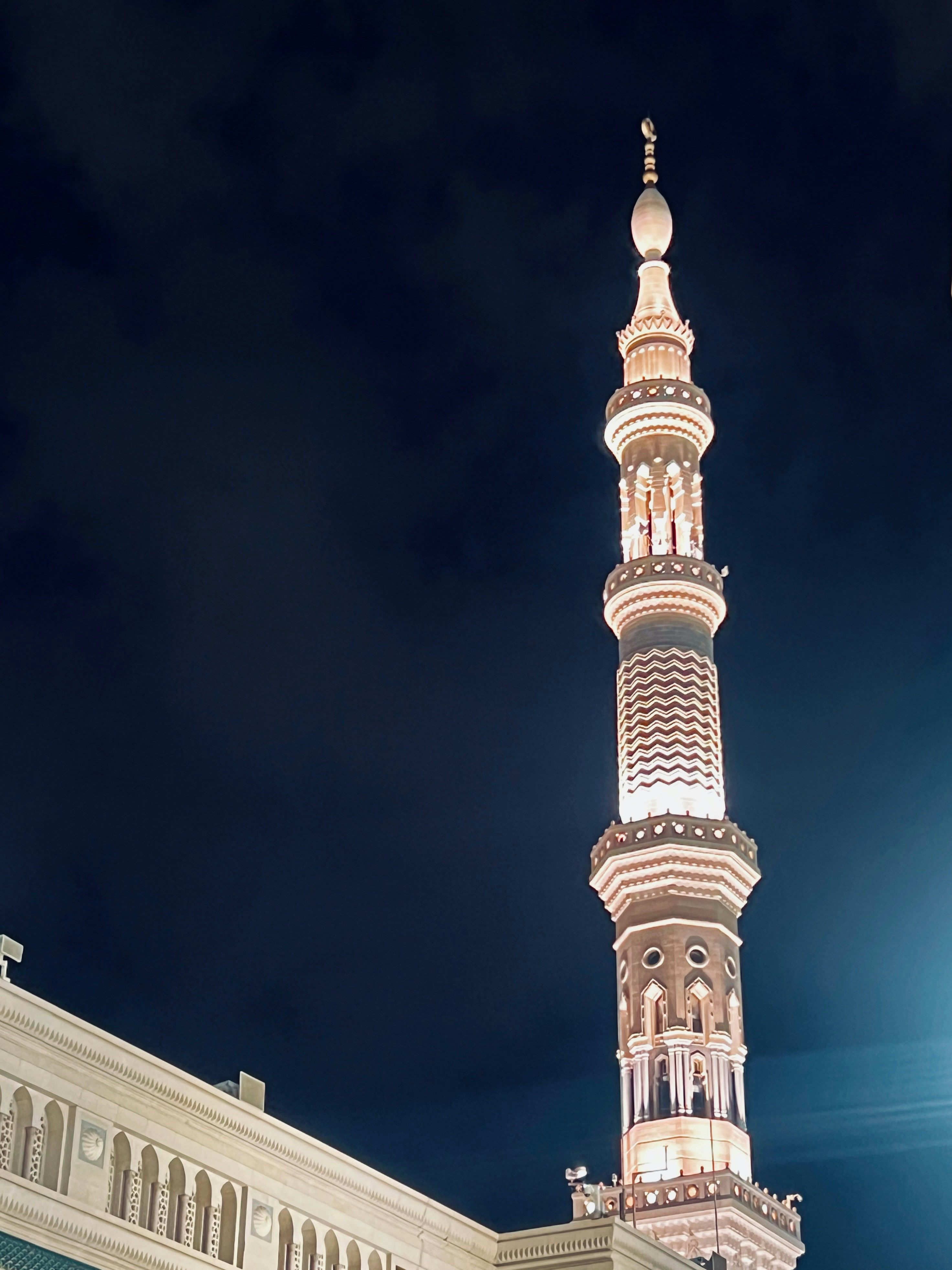 Tall illuminated minaret against a dark sky, with a portion of a white building at the base visible; architectural detail and nighttime lighting highlighted.