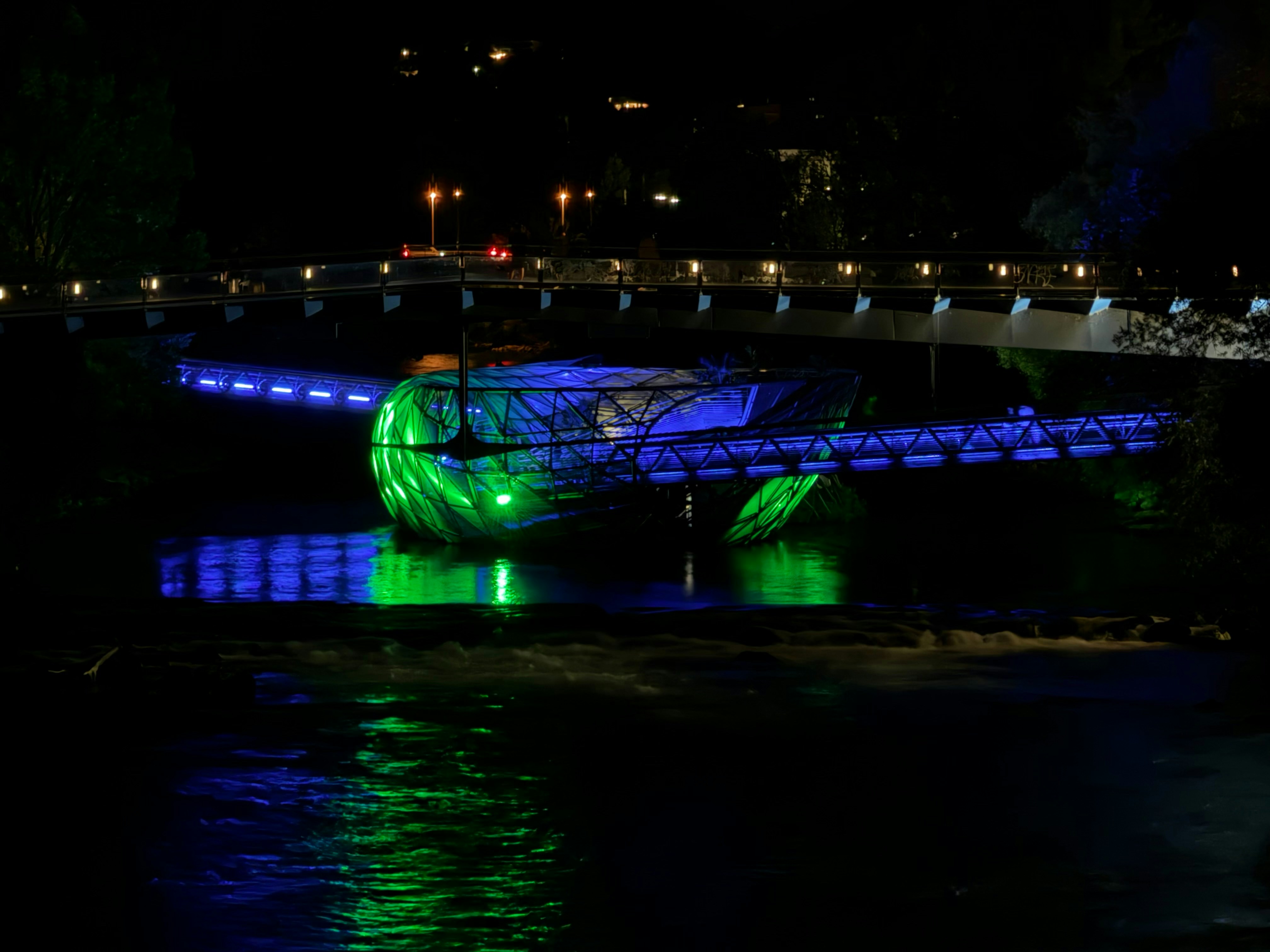 Neon-lit, aquatic sculpture glides beneath a dimly lit bridge, its green and blue hues reflecting on a quiet river at night. The scene emphasizes color and light interactions with still water.