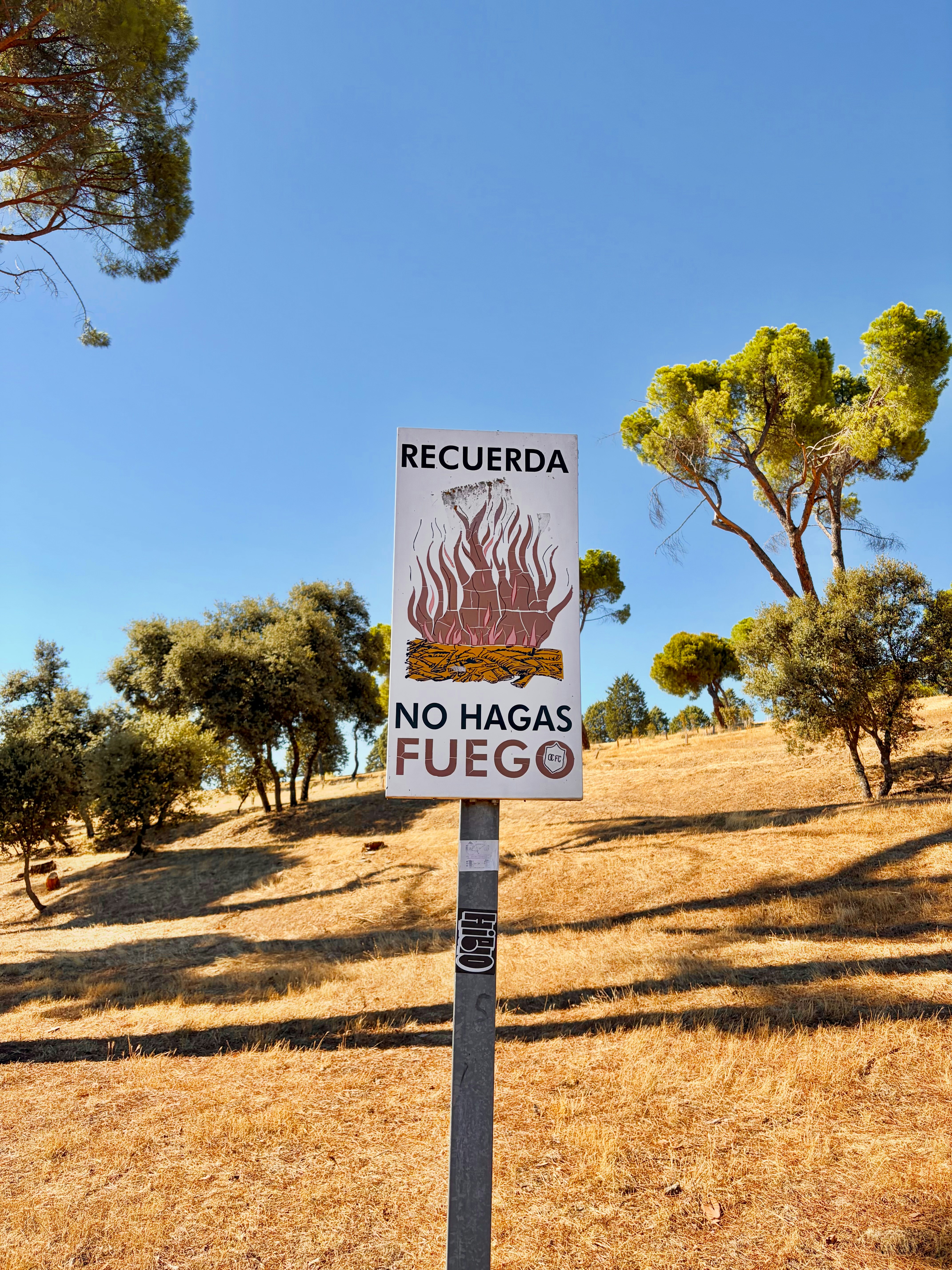 Bright sign on a post warning not to start fires, set against a dry hillside with scattered trees under a clear blue sky.
