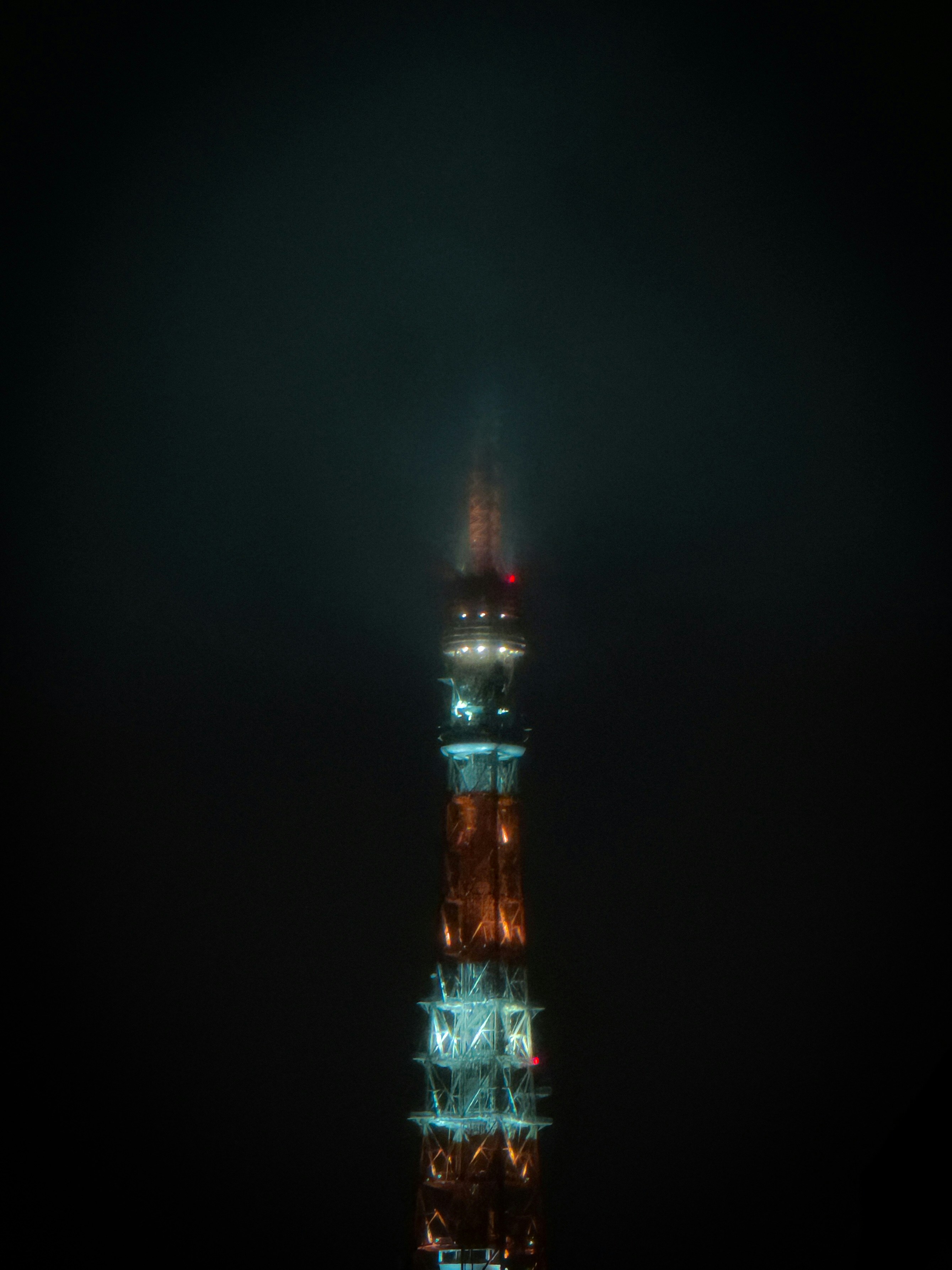 Illuminated broadcast tower rises from darkness, its structure bathed in cool blue and warm orange light. The scene emphasizes the tower’s vertical presence against a moody night sky.