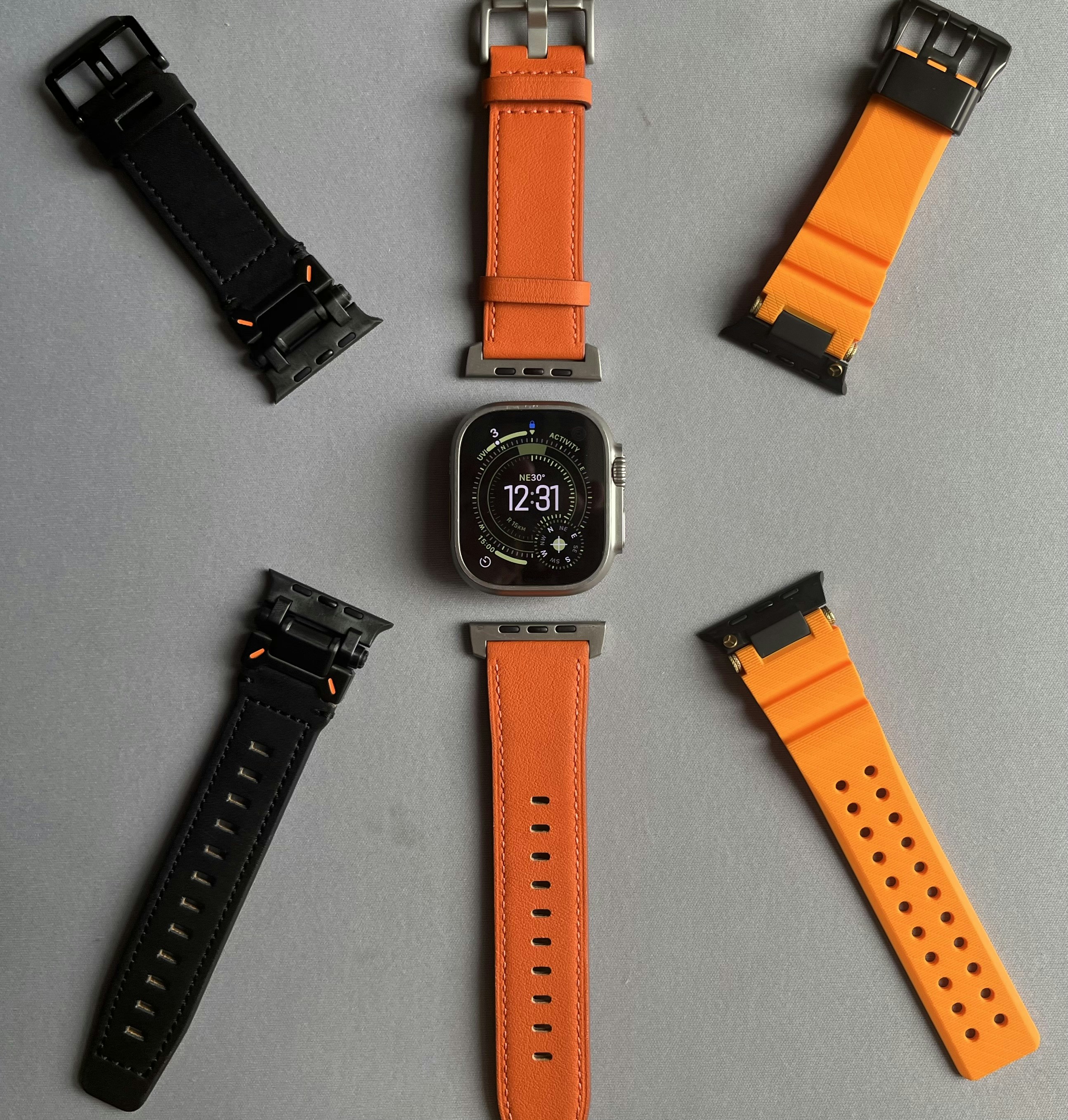 Multiple orange and black watch straps arranged in a circular pattern around a central smartwatch. The composition emphasizes color contrast and symmetry in a flat lay setup.