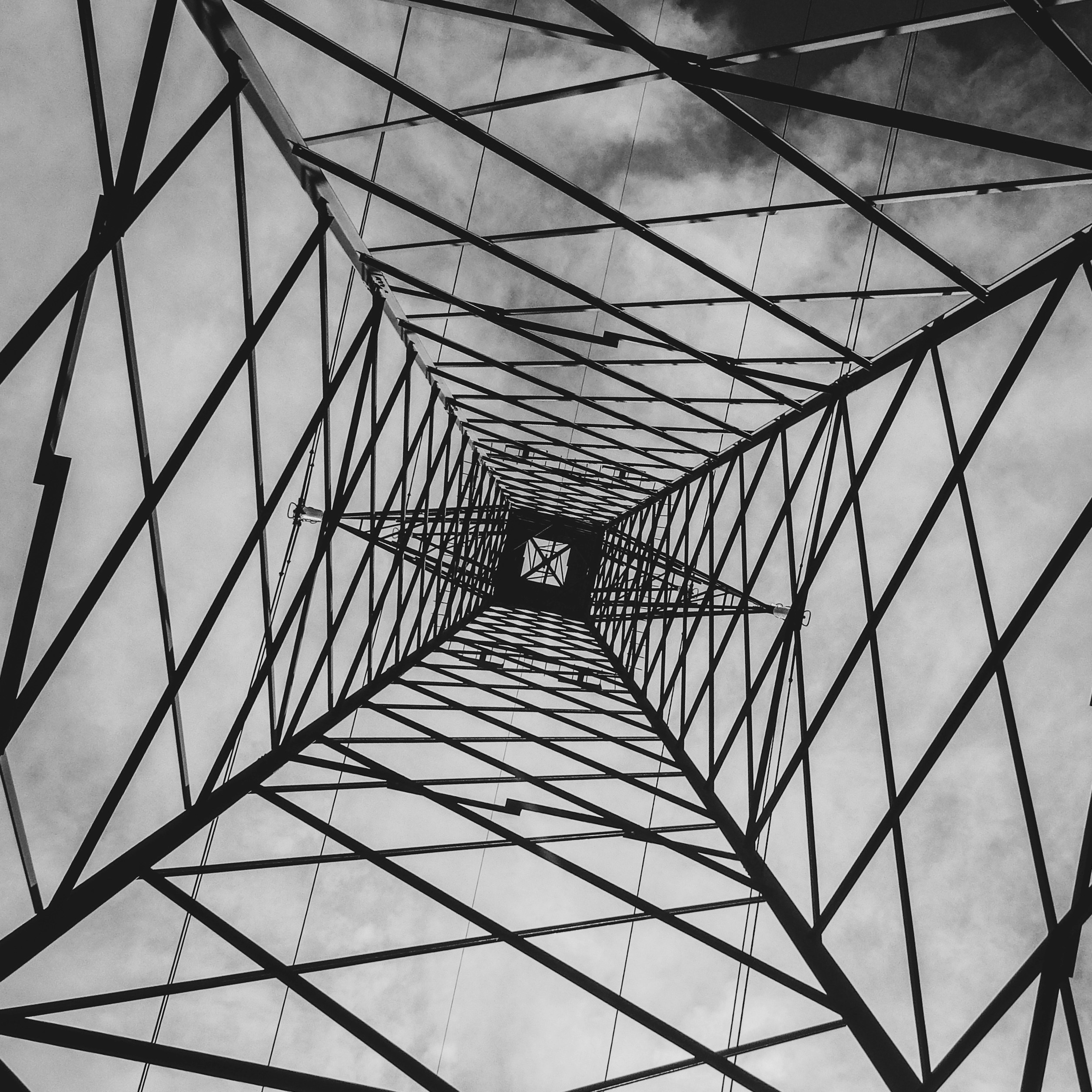 A direct low-angle view looking straight up through the steel frame of a transmission tower against the texture of the clouds.