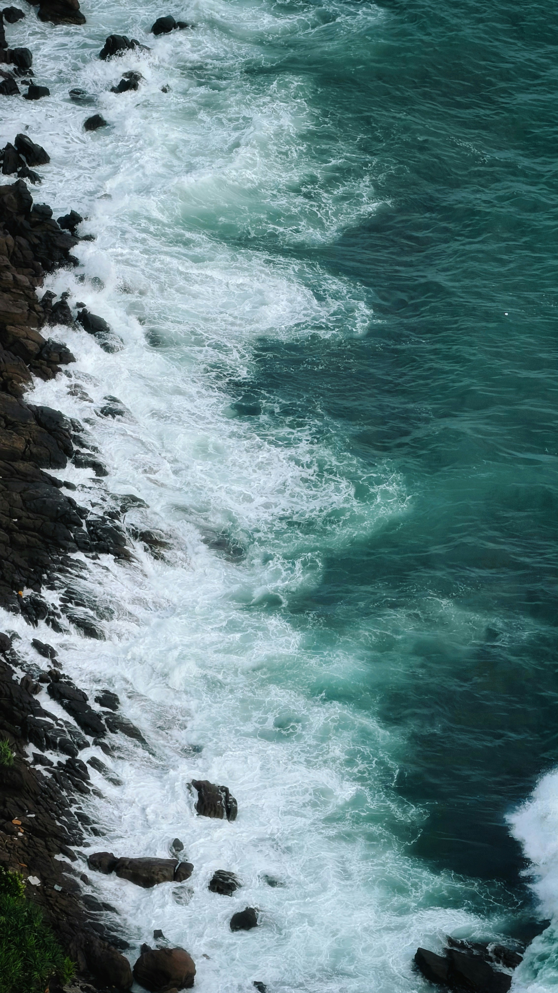 Coastal rocks meet turquoise surf with white foam along a steep shoreline. Aerial view emphasizes the contrast between dark basalt and teal water, highlighting the jagged coastline.