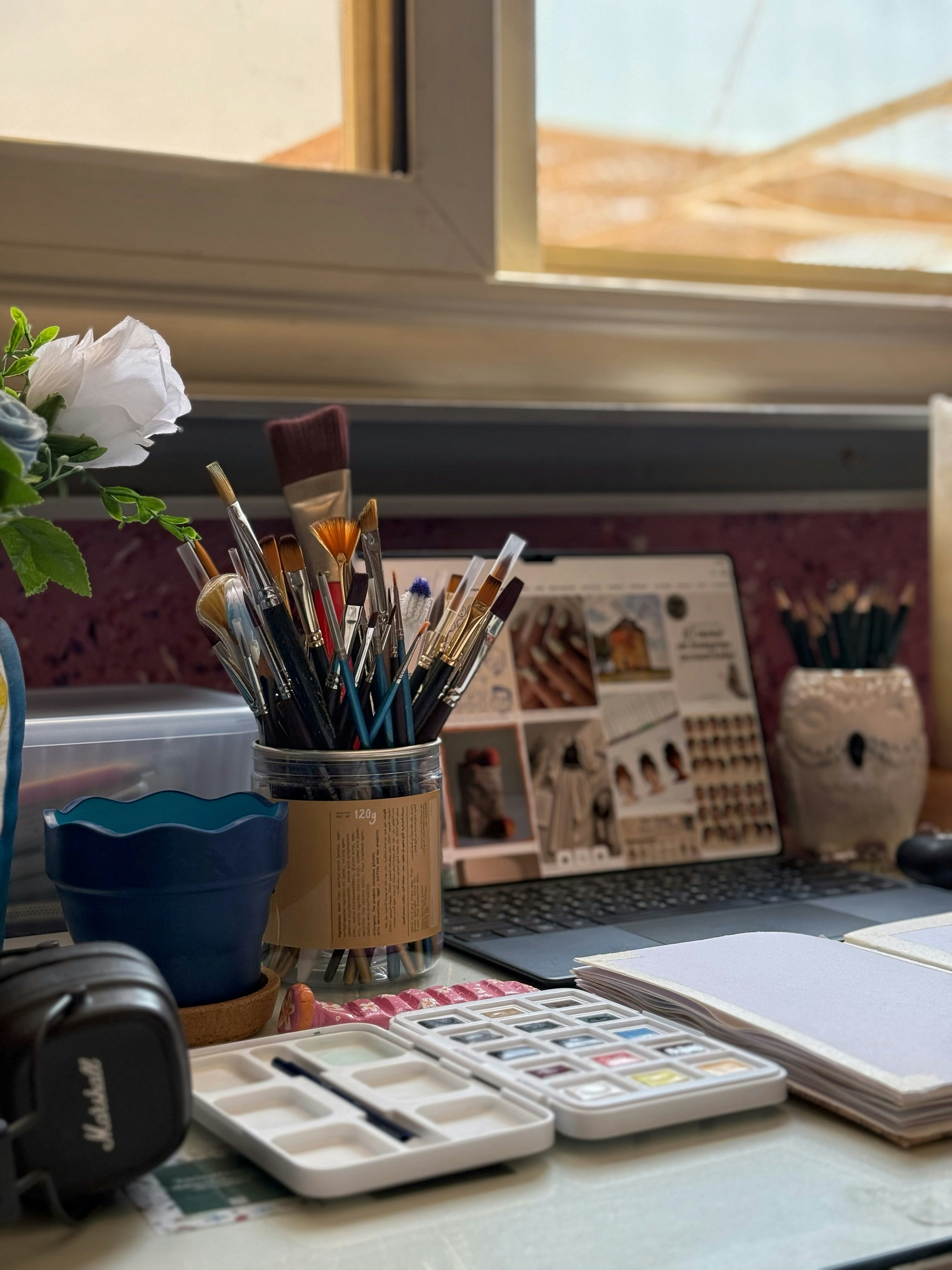 A creative workspace with a jar of paintbrushes, art supplies, and a laptop on a cluttered desk near a window. This is a photographic study of a craft area, showing tools and textures.